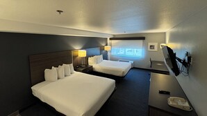 Quarto de hotel com duas camas, uma mesa, uma cadeira, uma TV e uma janela com persianas.