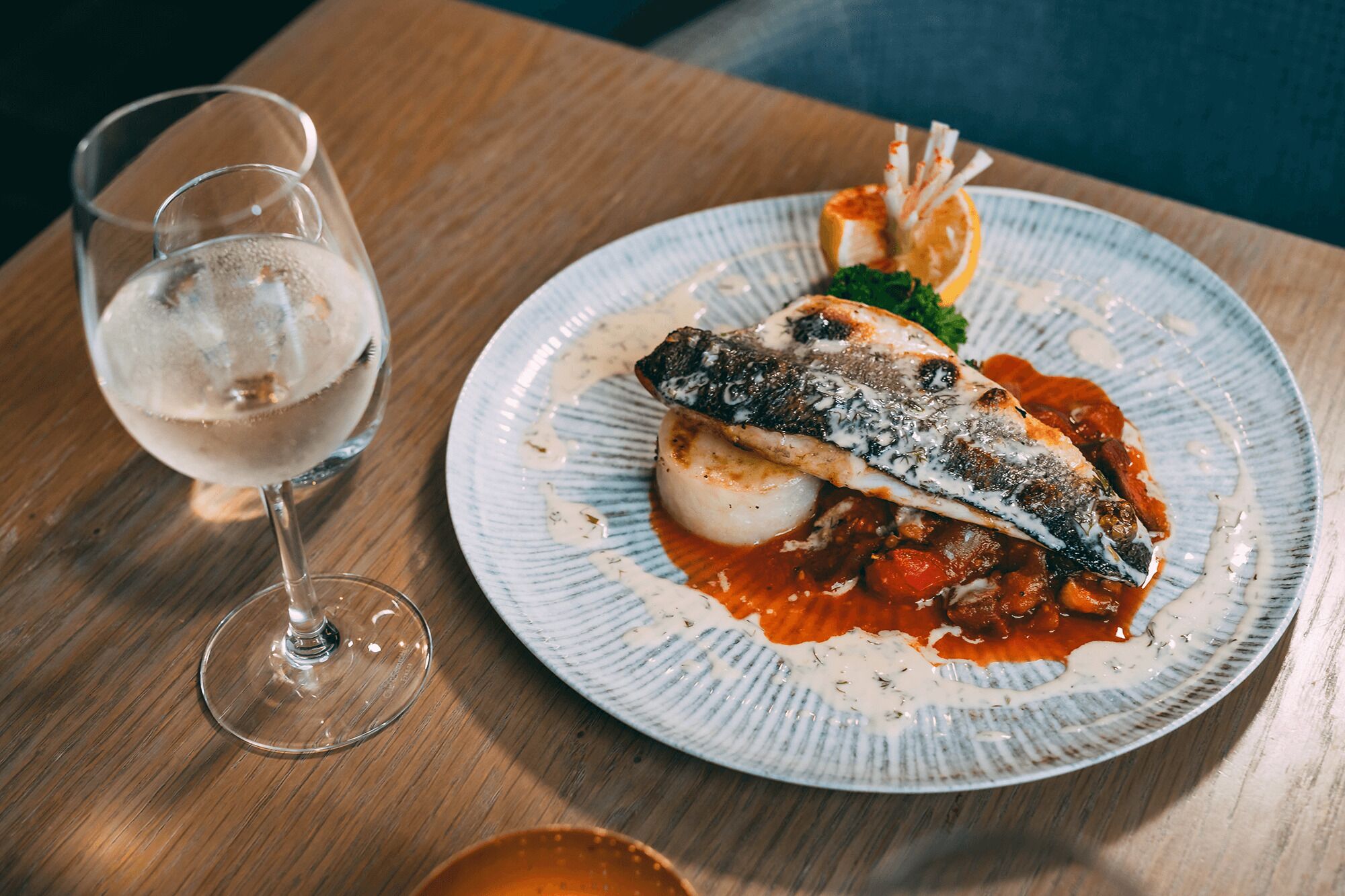 Prato de peixe com molho e legumes, um copo de vinho branco e uma mesa de madeira.