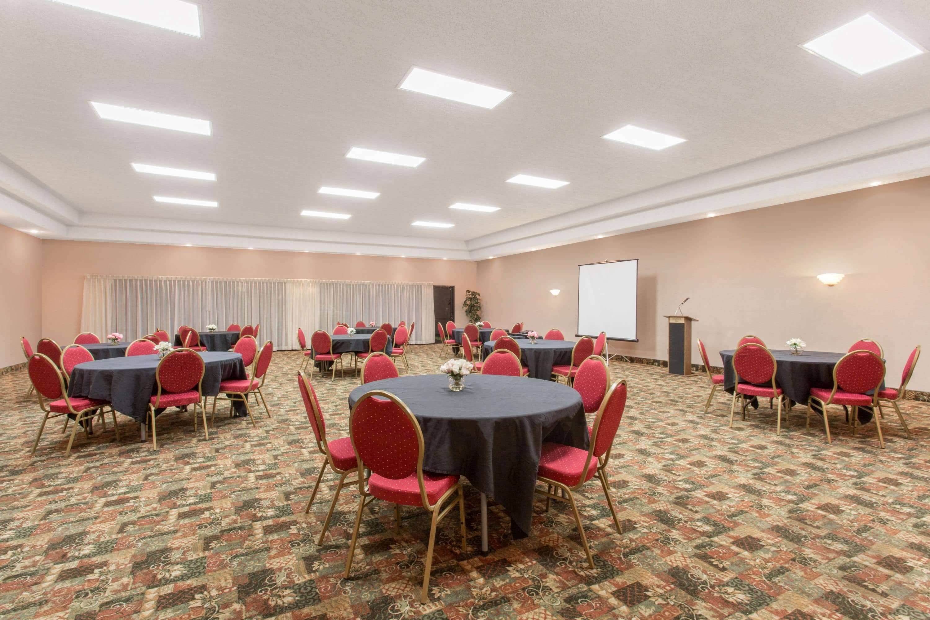 Sala de conferências com mesas redondas e cadeiras vermelhas, piso coberto por carpete e tela de projetor.