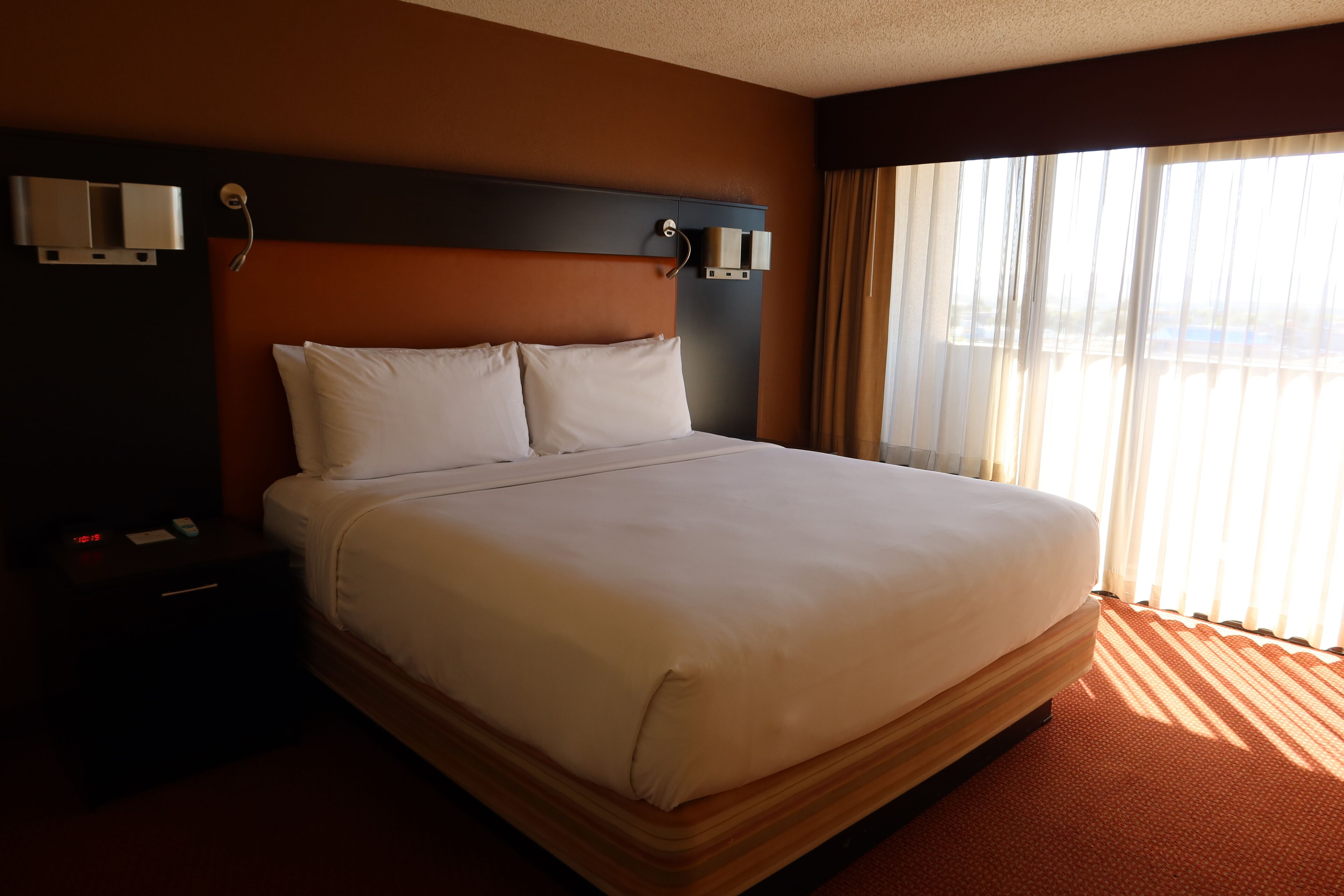 Quarto de hotel com uma cama grande, mesa de cabeceira e janela com cortinas.
