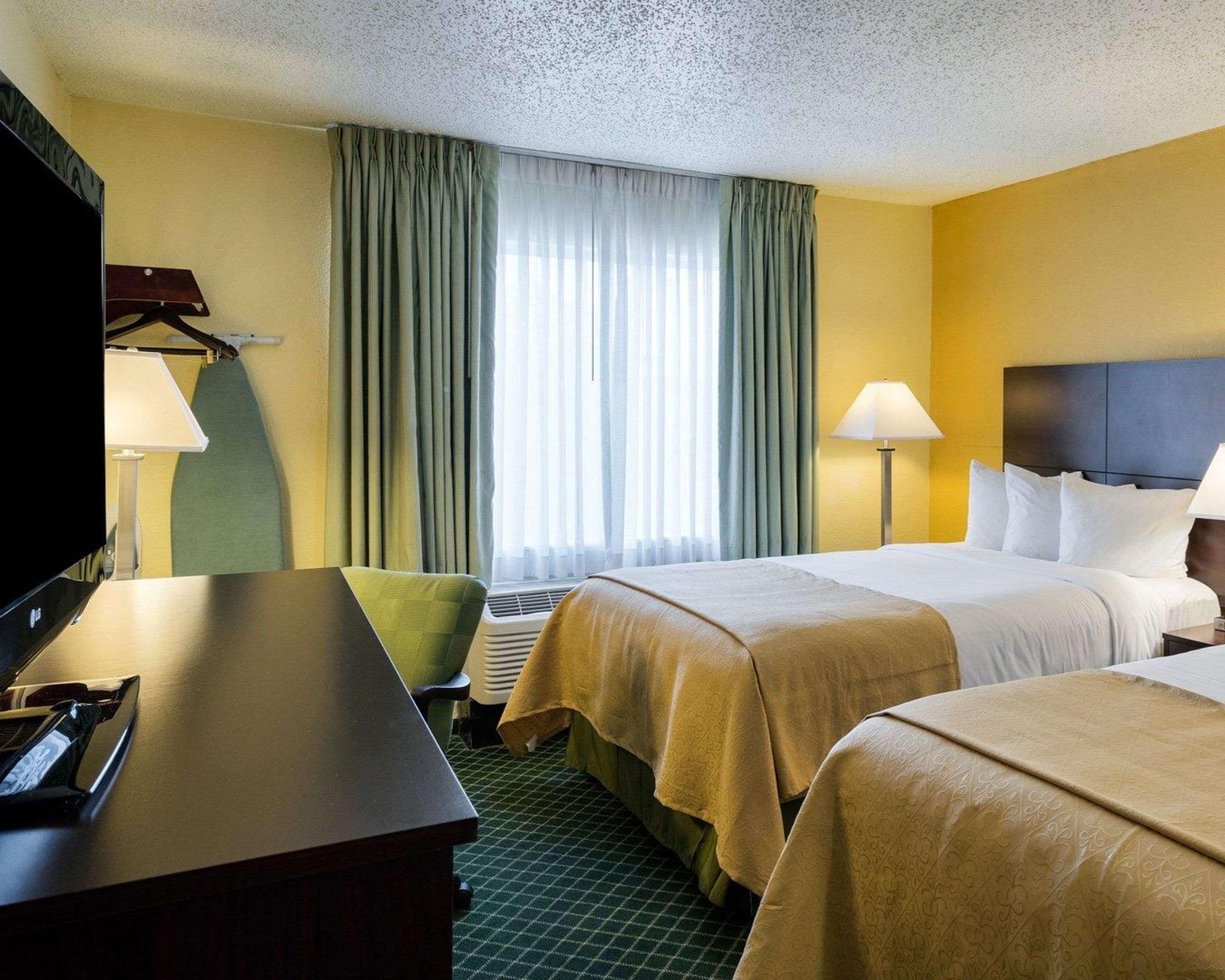 Quarto de hotel com duas camas, uma escrivaninha, televisão e janela com cortinas.