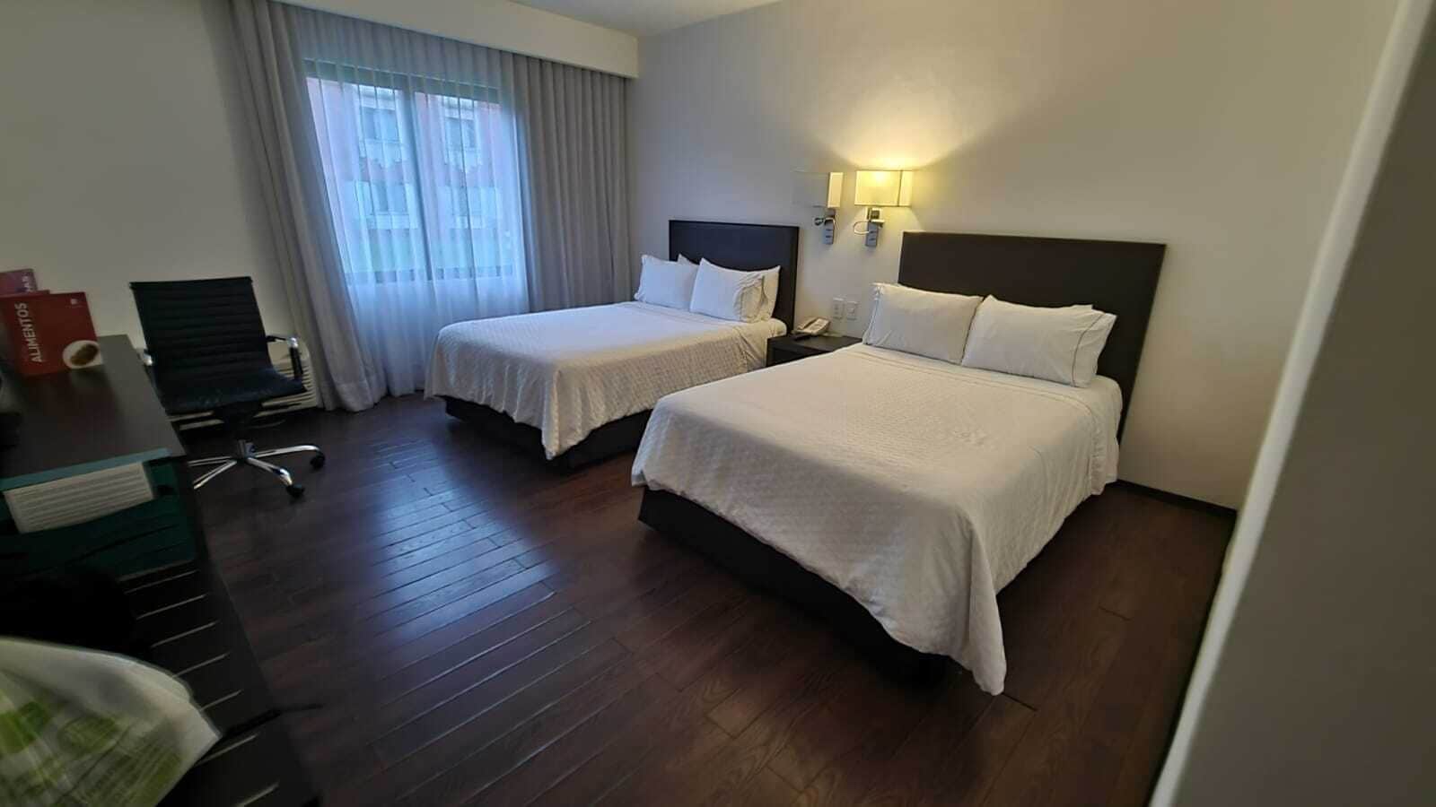 Quarto de hotel com duas camas, uma escrivaninha, uma cadeira e uma janela com cortinas.