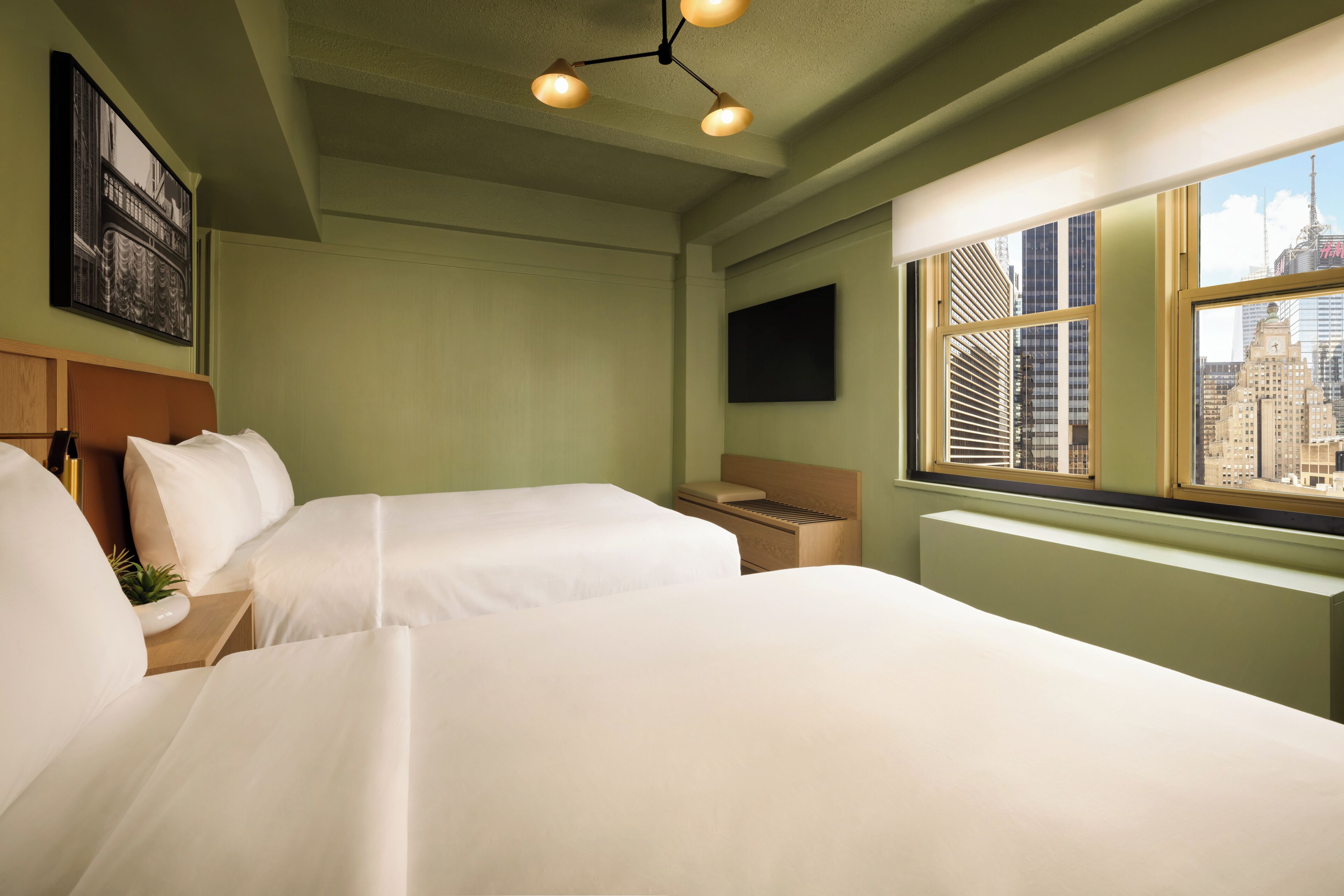 Quarto de hotel com duas camas, luminária no teto, televisão fixada na parede e janela com vista para a cidade.