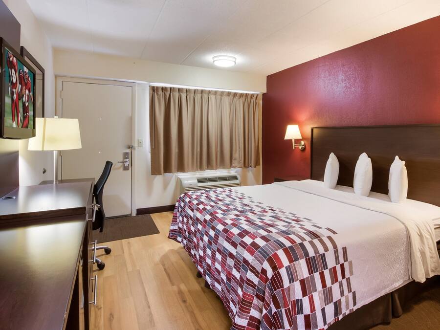 Quarto de hotel com cama, escrivaninha, cadeira, abajur, televisão fixada na parede e janela com cortinas.