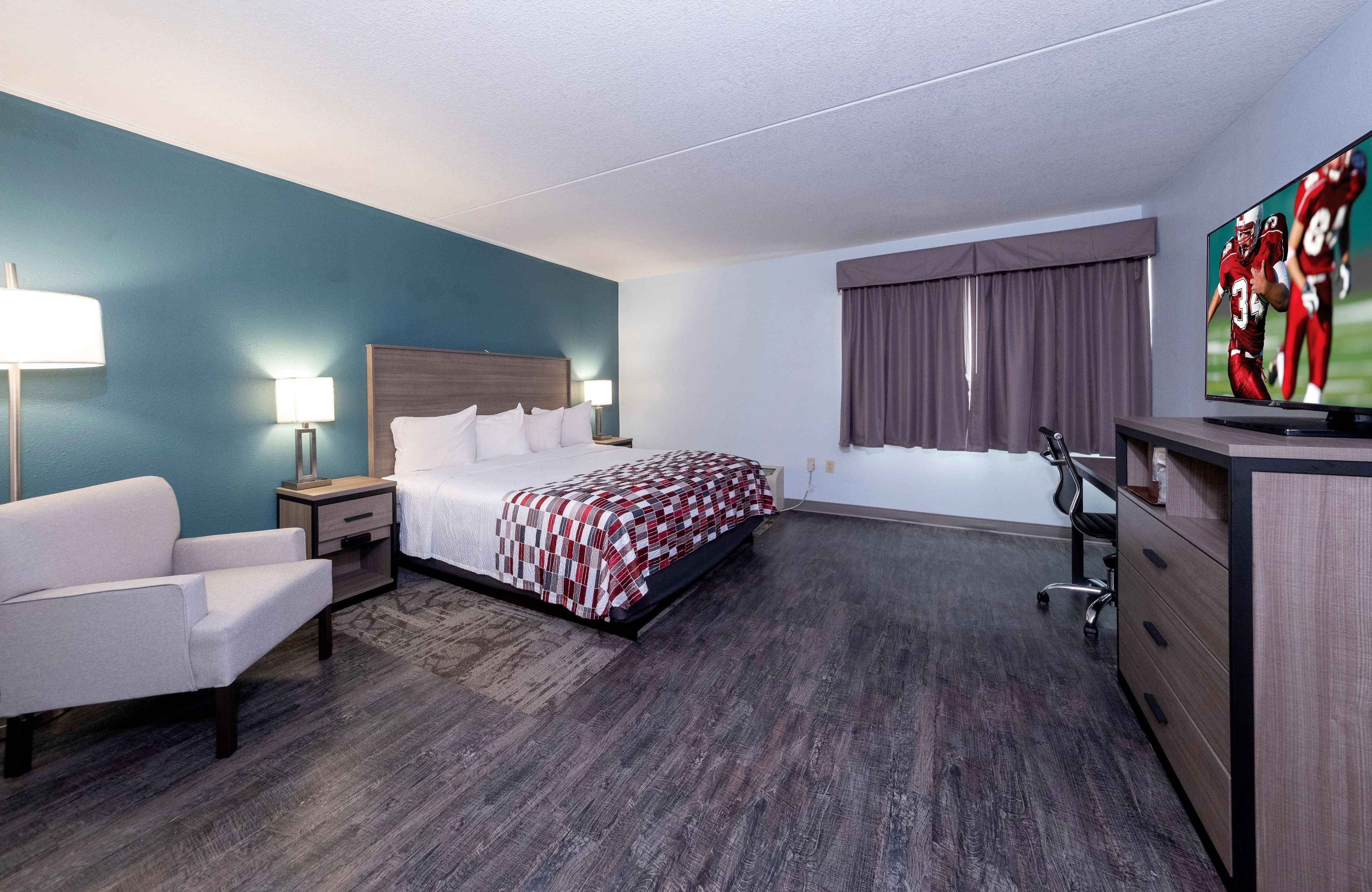 Quarto de hotel com cama, mesas de cabeceira, escrivaninha, cadeira, televisão e janela com cortinas.