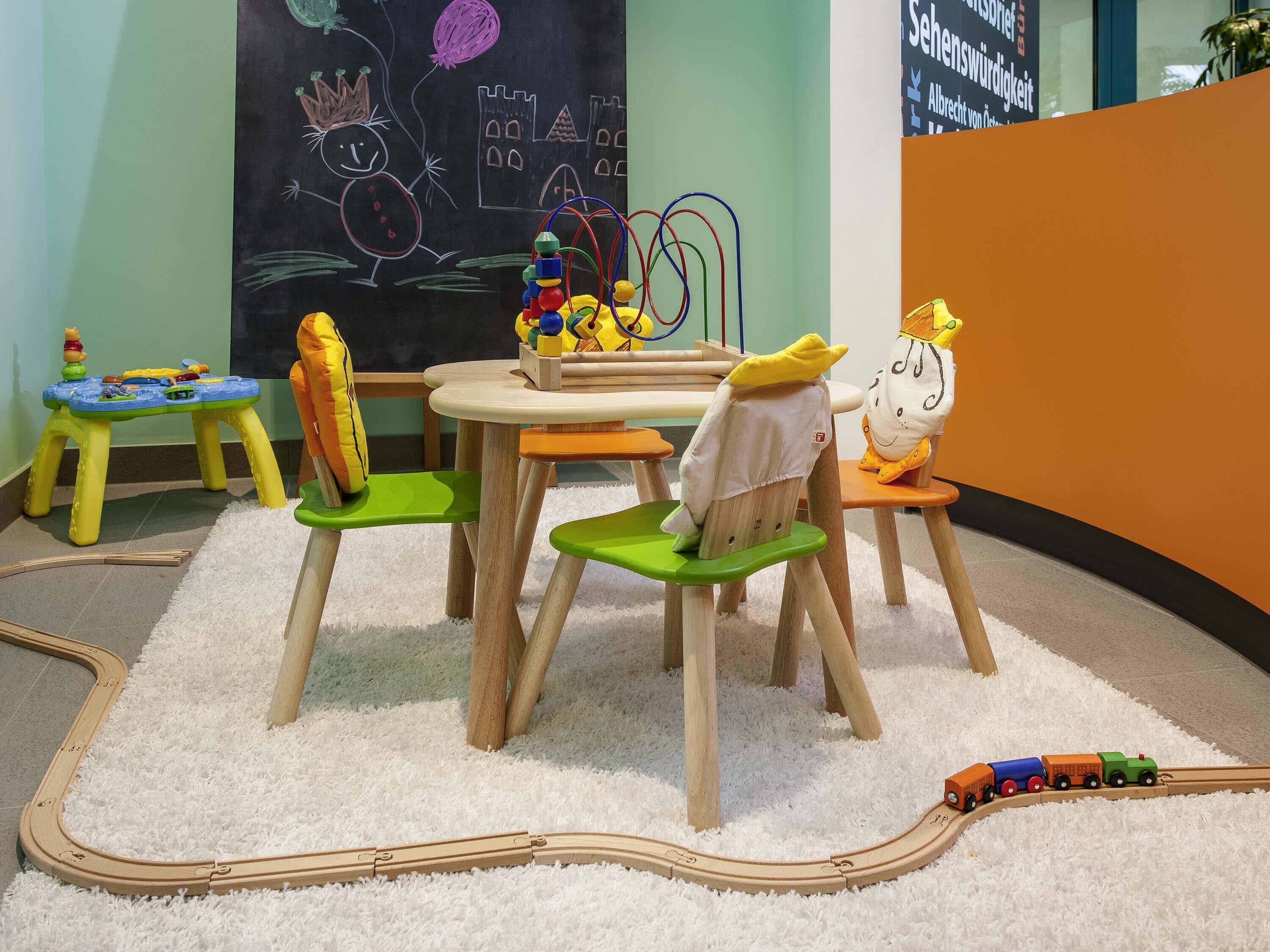 Uma área de recreação infantil com uma mesa de madeira, cadeiras, uma pista de trem de brinquedo e um quadro negro com desenhos.