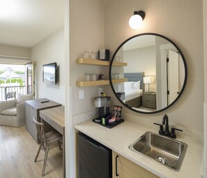 Quarto de hotel compacto com cozinha compacta, cama com encosto, espelho redondo e pequena área de estar com uma cadeira e uma mesa.