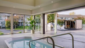 Uma área moderna de piscina interna com paredes de vidro transparente, vista para o exterior e um pequeno espaço de estar ao ar livre.