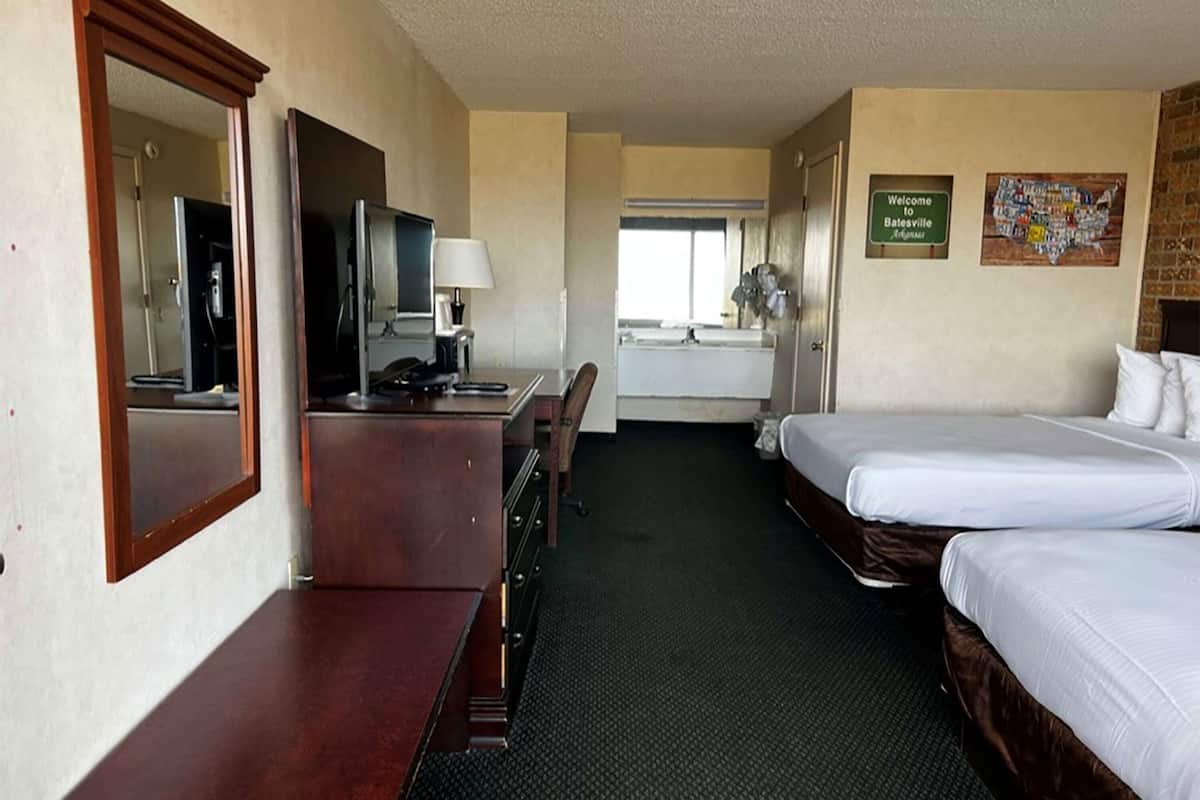 Quarto de hotel com escrivaninha, duas camas, espelho, televisão e janela.