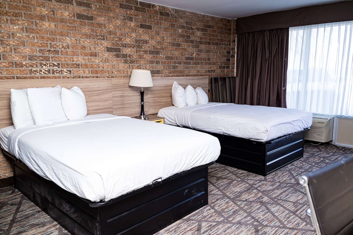 Quarto de hotel com duas camas, uma parede de tijolos, uma janela grande com cortinas e um tapete estampado.