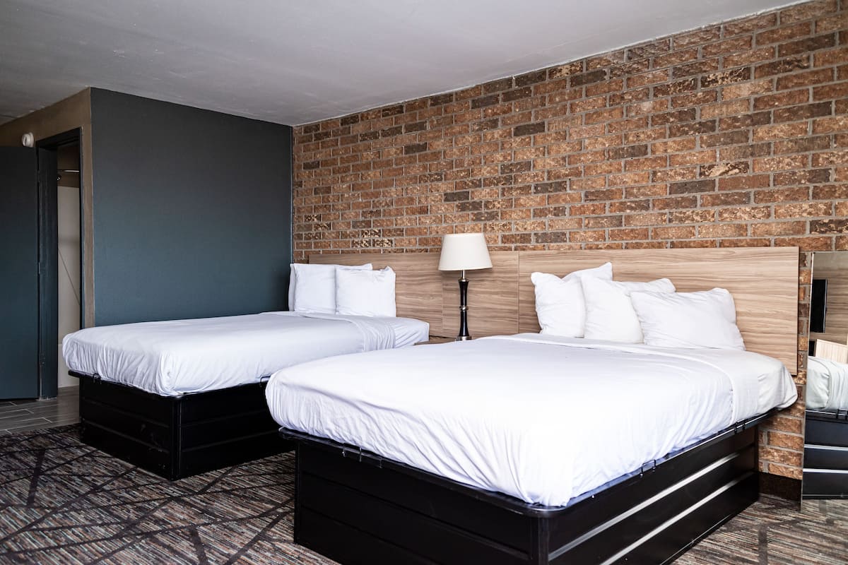 Quarto de hotel com duas camas de solteiro, parede de tijolos, cabeceira de madeira e carpete estampado.