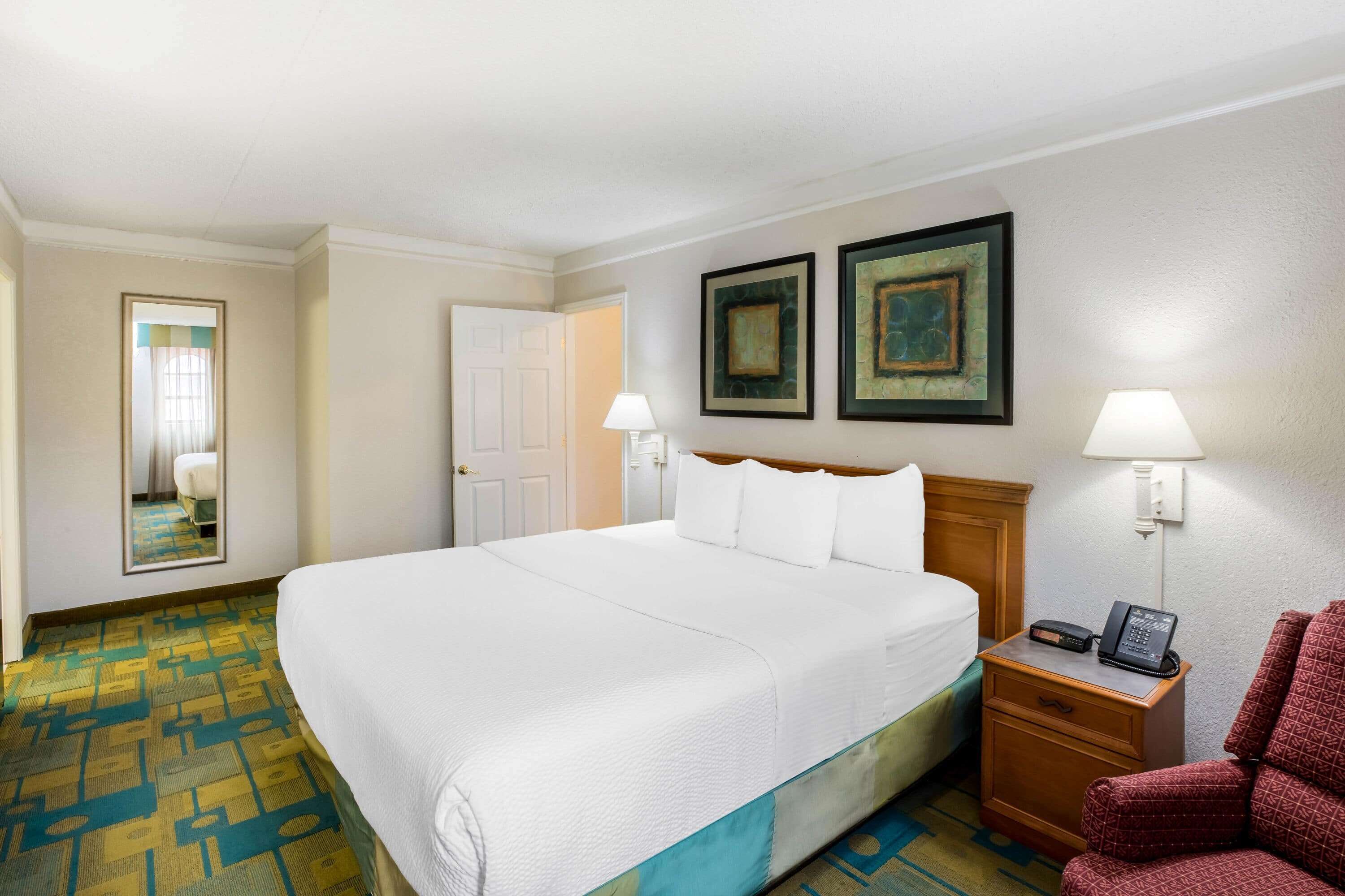 Quarto de hotel com uma cama grande, uma mesa de cabeceira com telefone, uma cadeira e duas fotos emolduradas na parede.