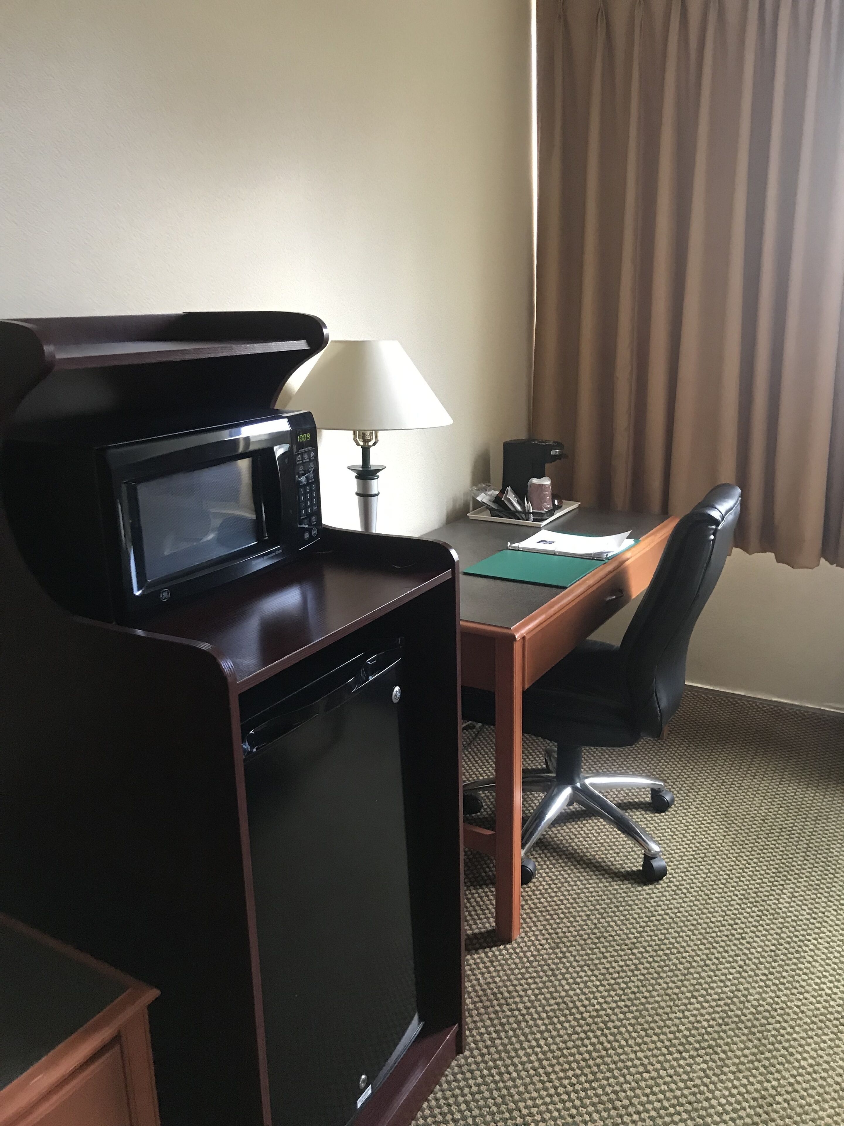 Quarto de hotel compacto com cozinha compacta, escrivaninha, cadeira, luminária e janela com cortinas.
