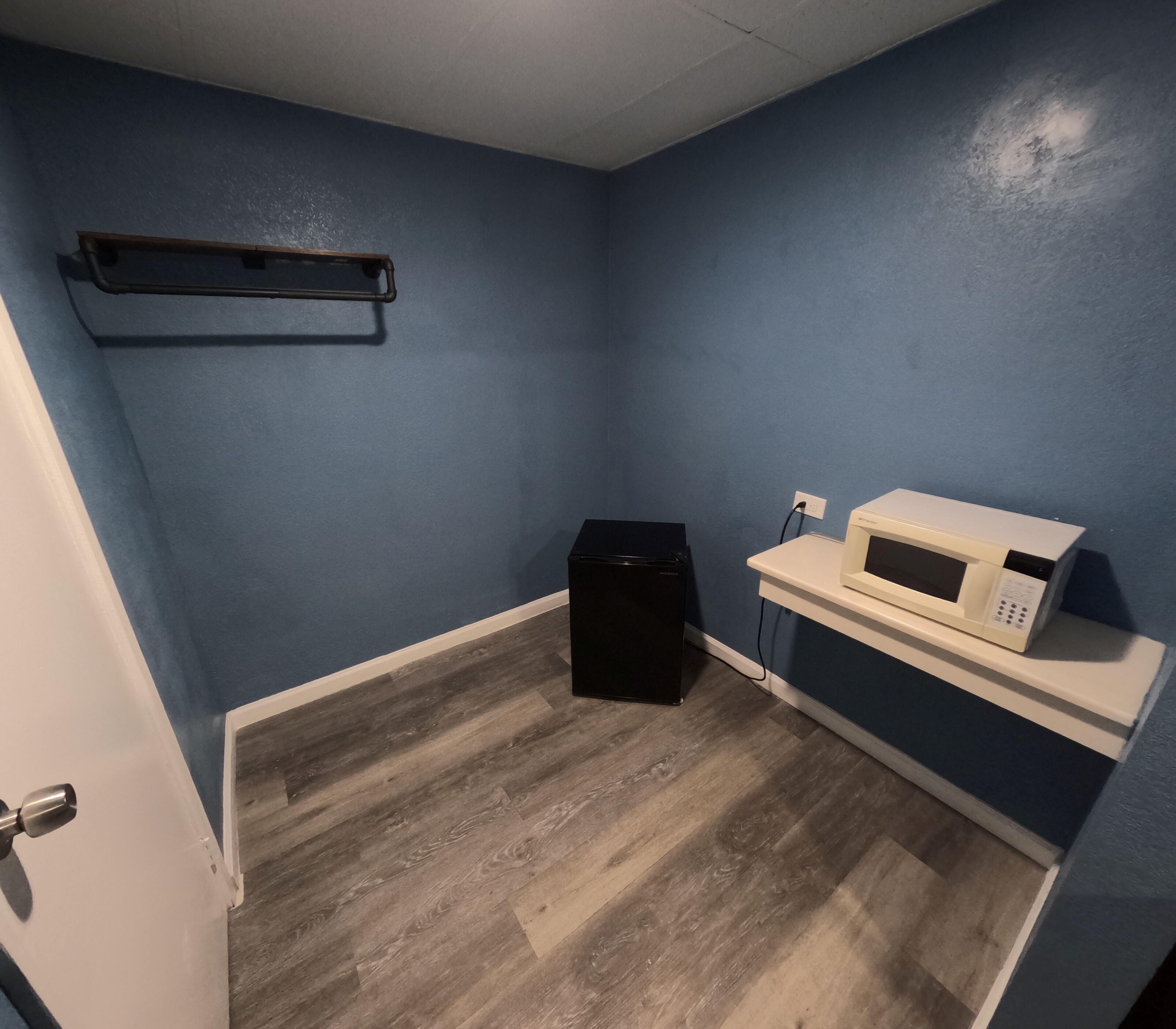 Um quarto pequeno com paredes azuis, chão de madeira, um cesto de lixo preto e um micro-ondas numa prateleira branca.