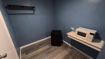 Um quarto pequeno com paredes azuis, chão de madeira, um cesto de lixo preto e um micro-ondas numa prateleira branca.