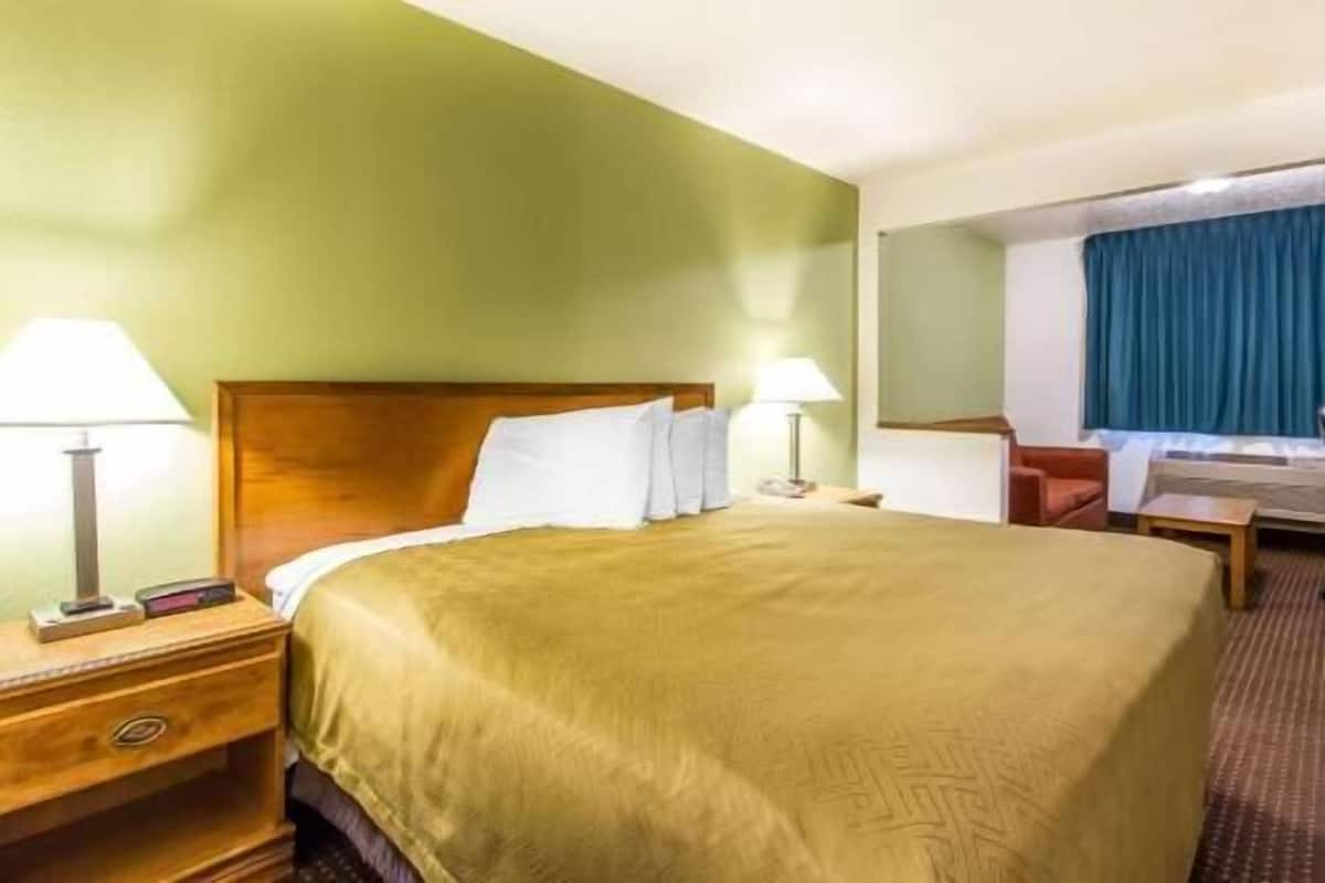 Quarto de hotel com cama, mesas de cabeceira, luminária, cadeira e janela com cortinas.