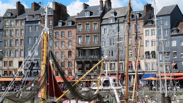 Uma área histórica à beira-mar, com edifícios tradicionais europeus, um navio à vela imponente e uma movimentada cena portuária.