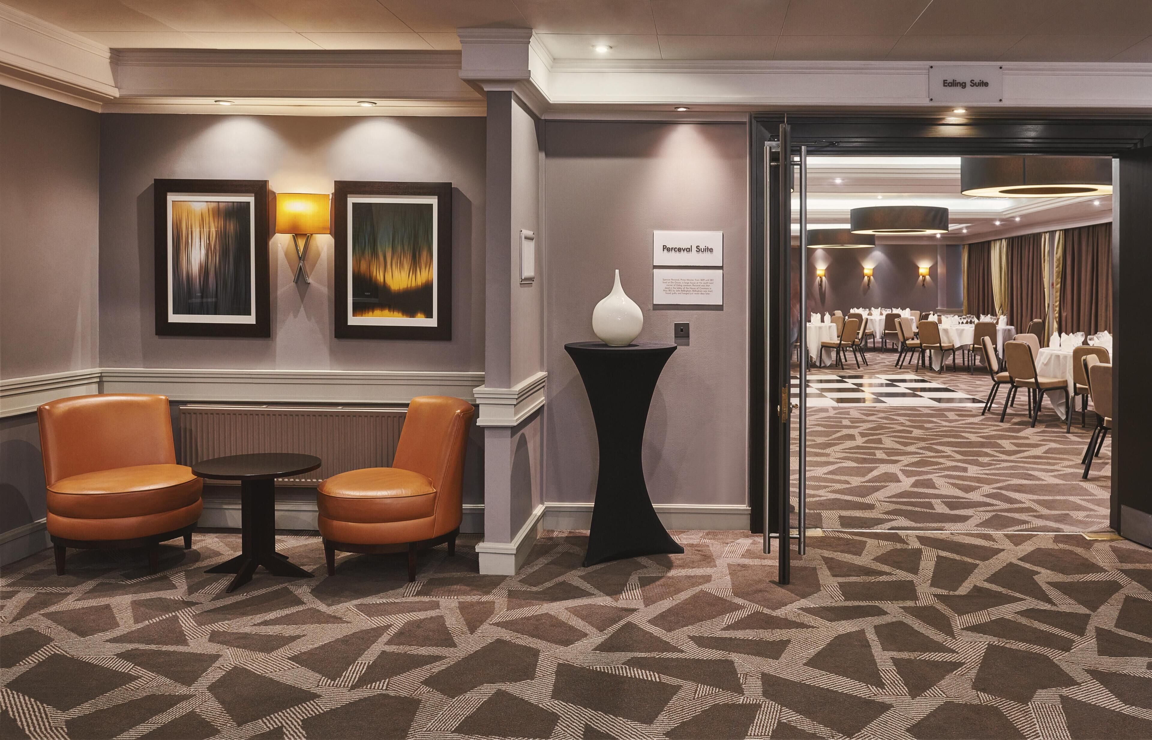 Um saguão de hotel com uma mesa de recepção, duas poltronas de couro, uma mesa pequena, obras de arte emolduradas e uma placa indicando a Suíte Perceval.