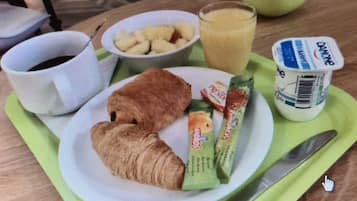 Mesa de café da manhã com croissants, uma tigela de frutas, um copo de suco e um pote de iogurte.