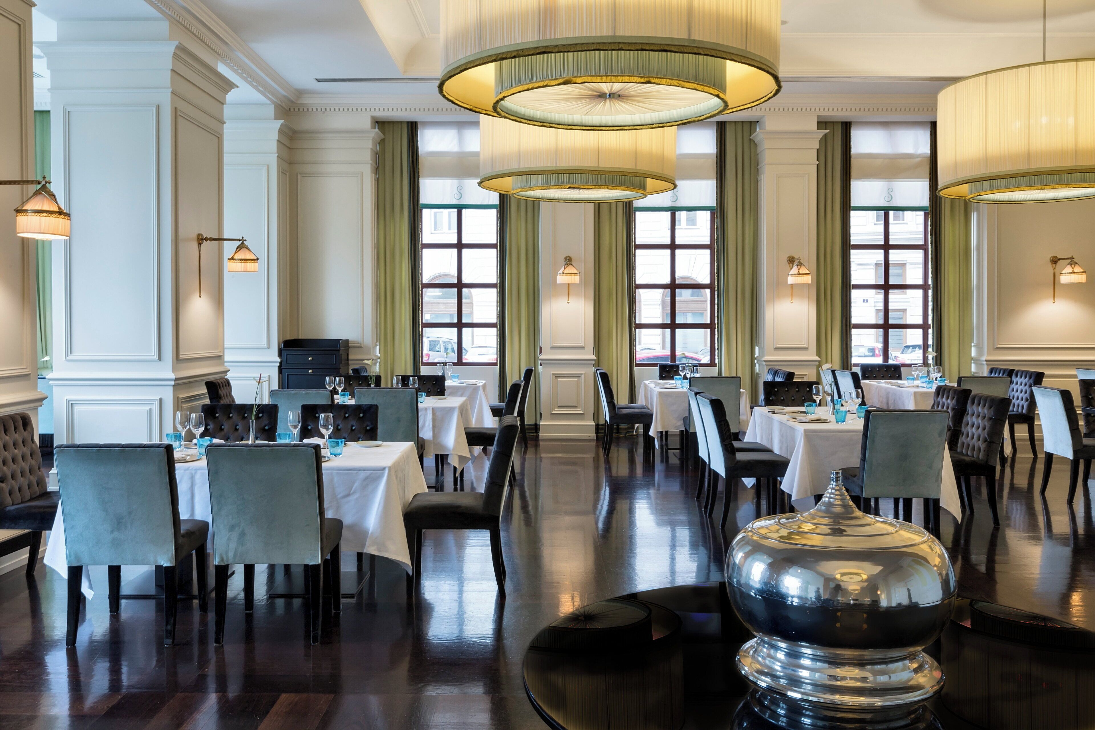 Uma sala de jantar formal com várias mesas e cadeiras, janelas amplas e luminárias elegantes.