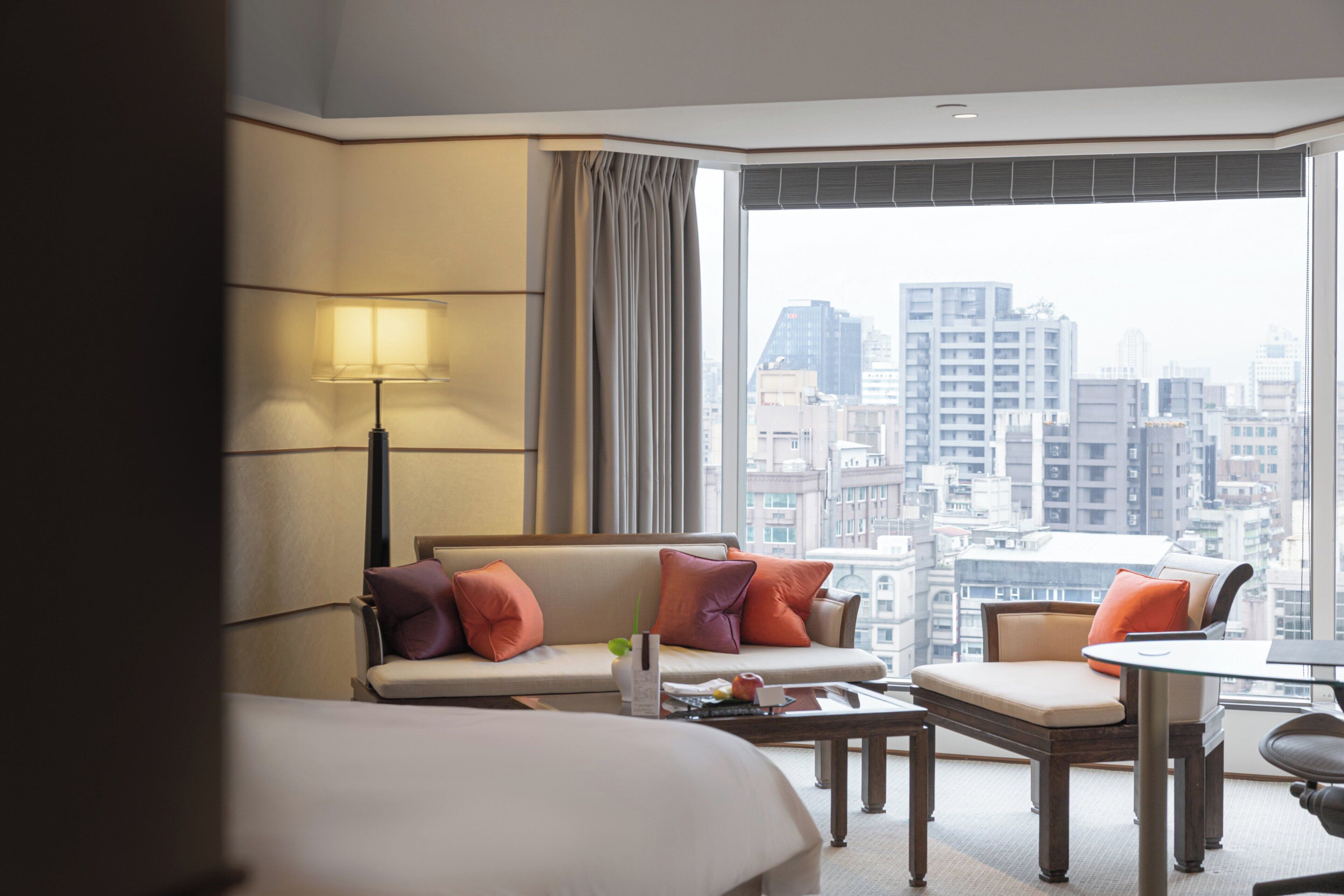 Quarto de hotel com uma janela grande, um sofá, uma cadeira, uma mesa pequena e um abajur.