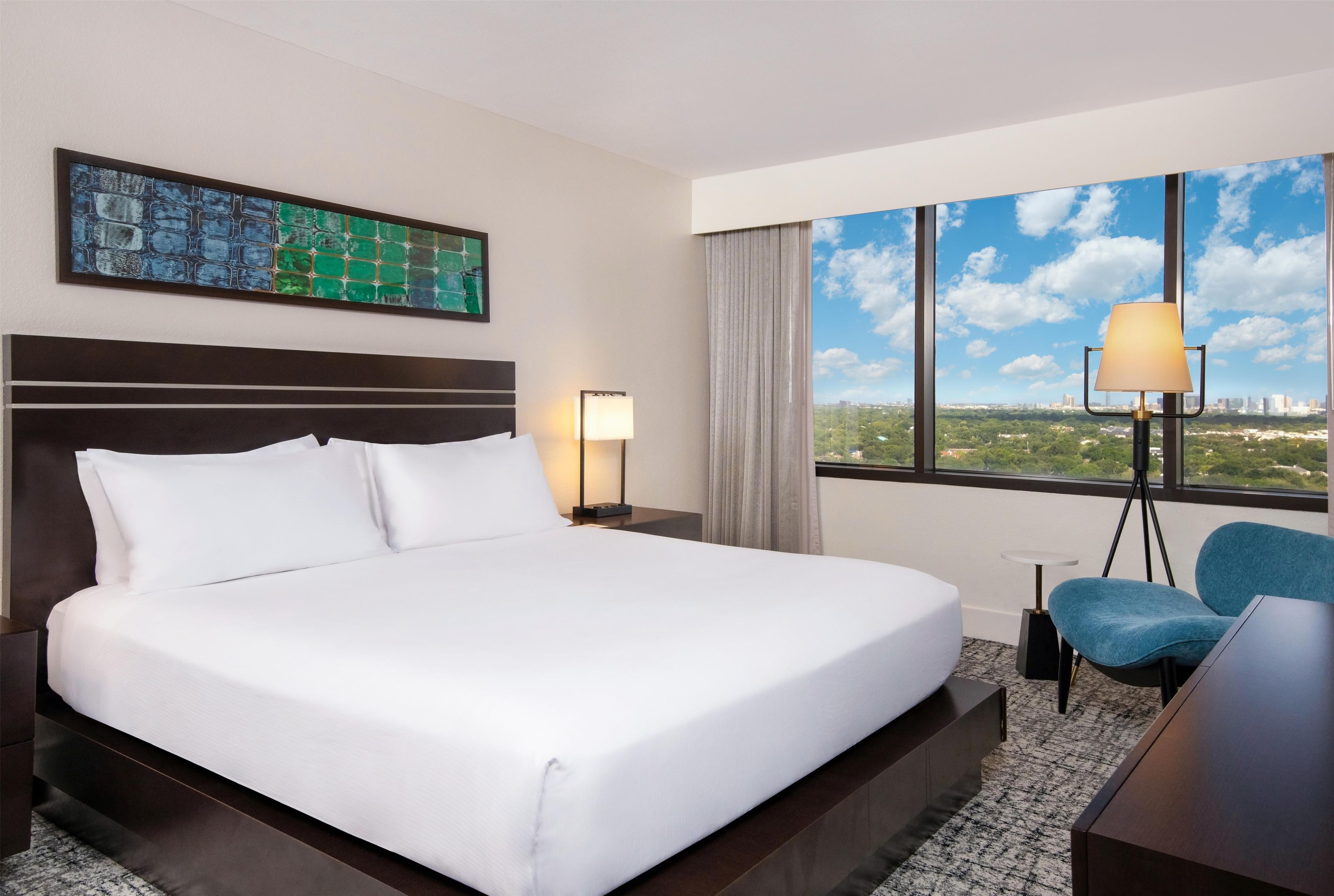 Quarto de hotel com cama grande, escrivaninha, cadeira, luminária, vista da cidade e um quadro na parede.