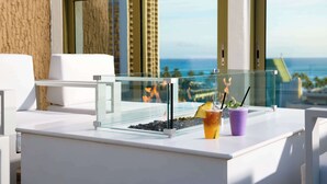 Uma varanda com divisória de vidro, uma pequena mesa com bebidas e vista para o oceano e edifícios.