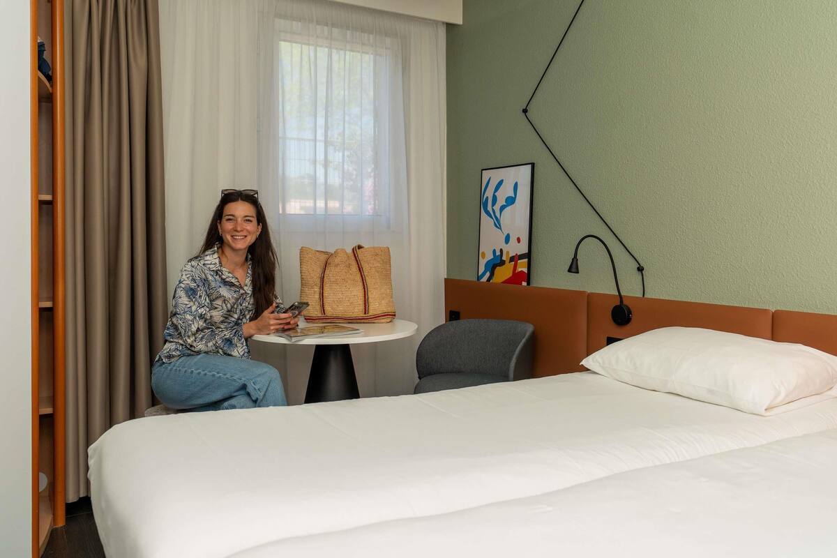 Quarto de hotel com cama, mesinha redonda, cadeira, abajur e janela com cortinas.