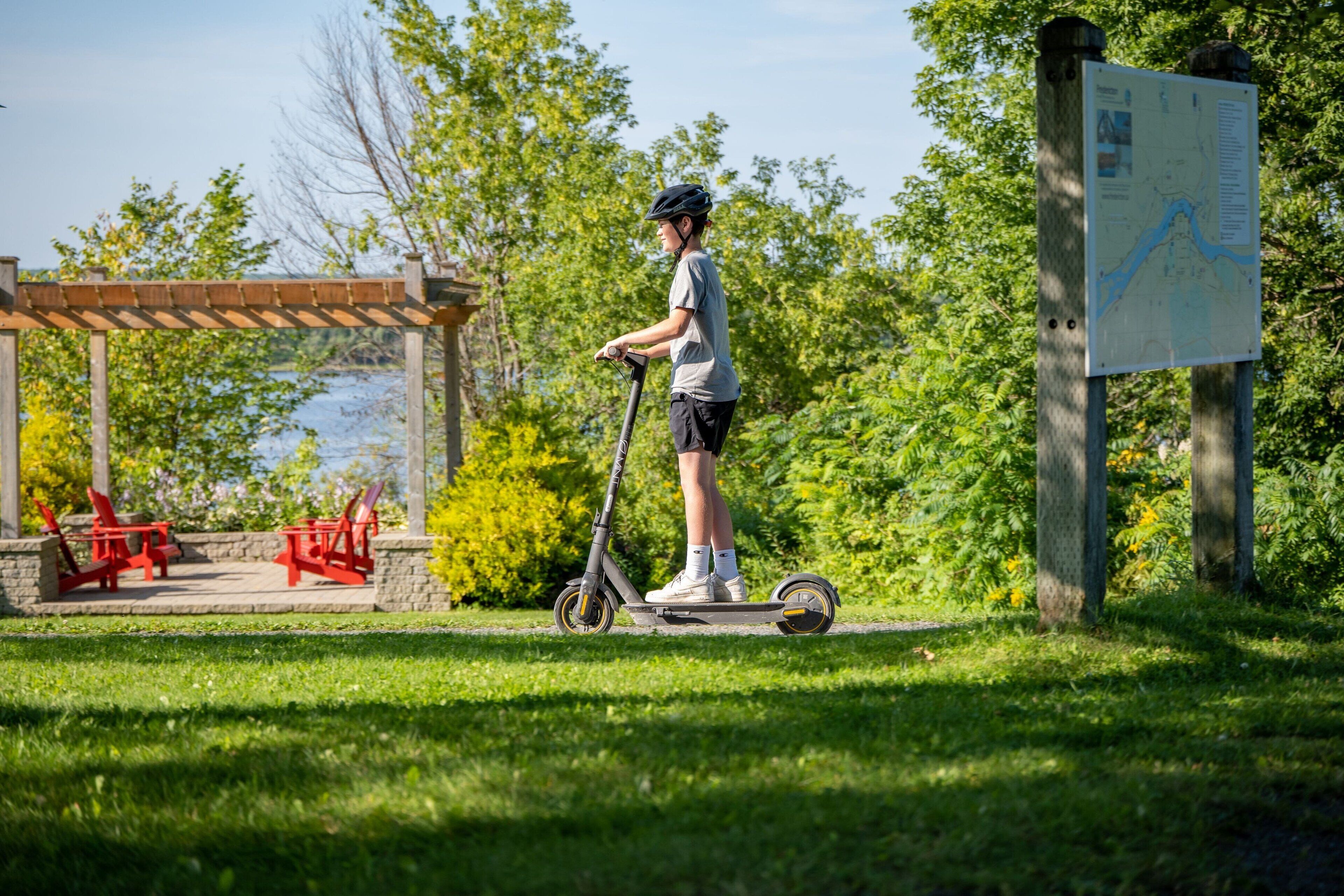 Uma pessoa andando de scooter por um caminho pavimentado em um parque, com uma pérgola de madeira e cadeiras vermelhas ao fundo.