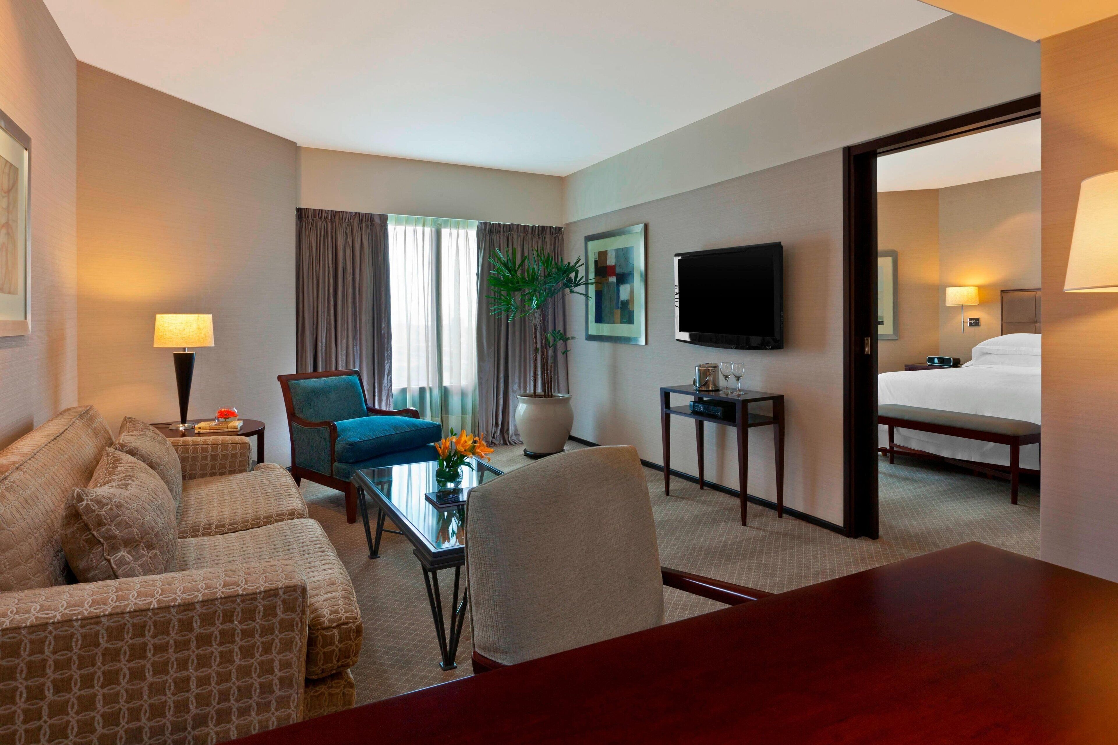 Quarto de hotel com sofá, uma cadeira, mesa de centro com flores, televisão, mesa de cabeceira e cama.