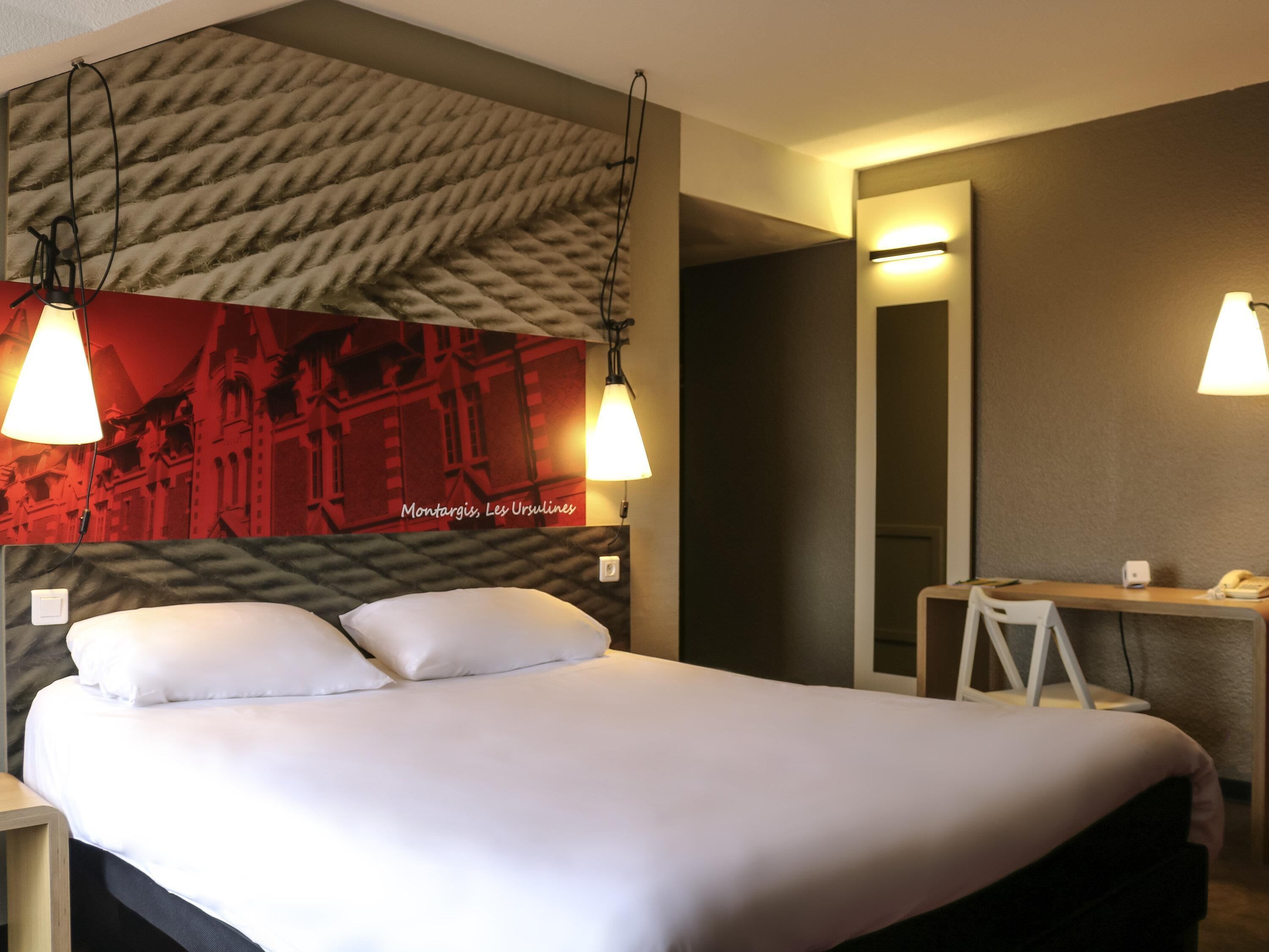 Quarto de hotel com cama, mesa de cabeceira, escrivaninha e espelho.