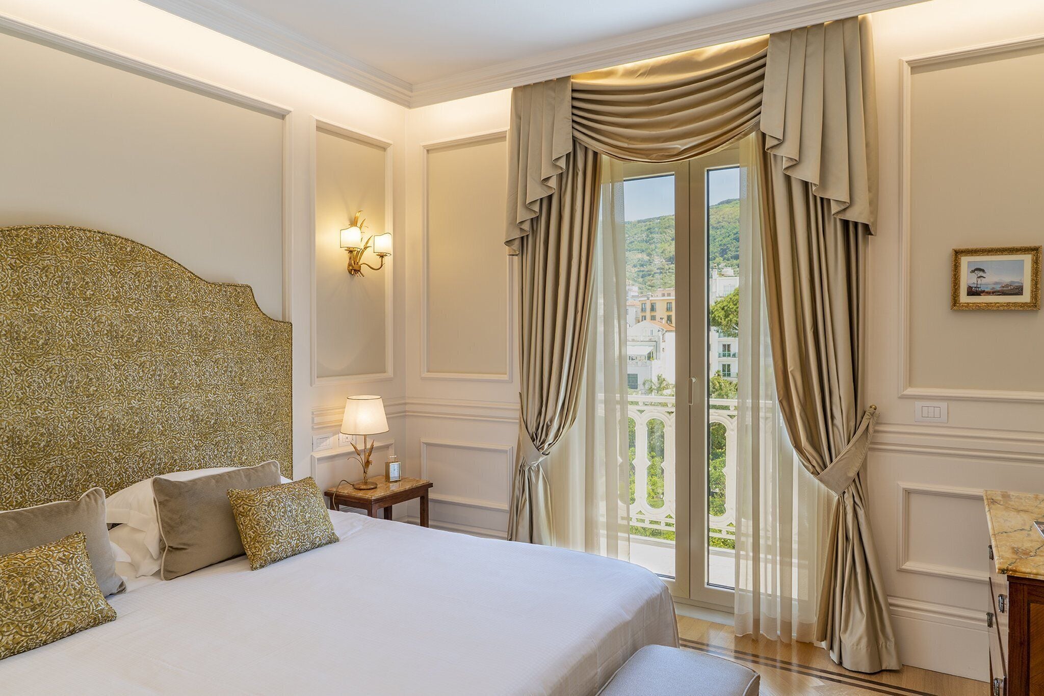 Quarto de hotel com uma cama grande, uma mesa de cabeceira com abajur e vista para uma varanda com cortinas.
