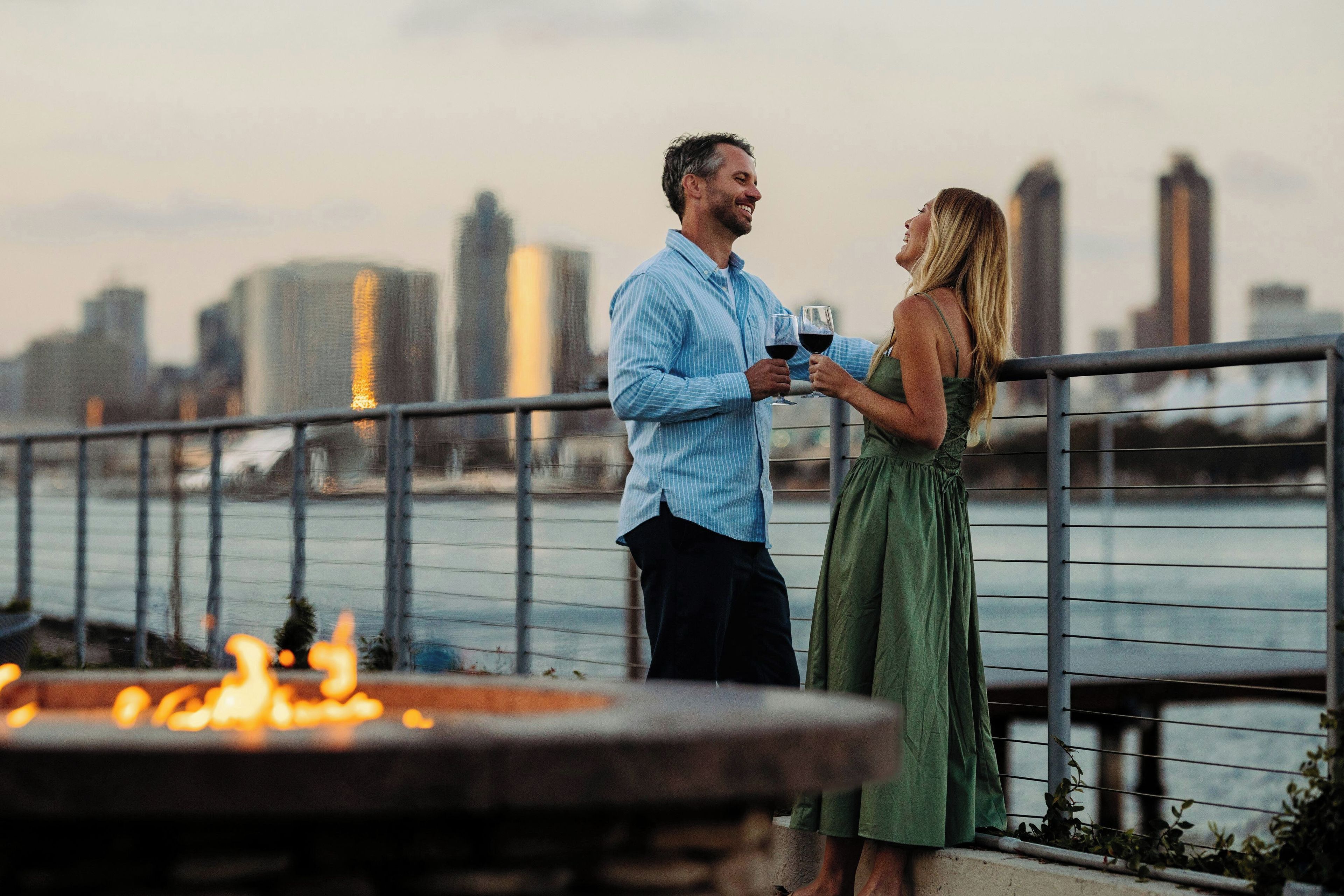 Um casal em pé perto de um corrimão, com uma vista da linha do horizonte da cidade ao fundo.