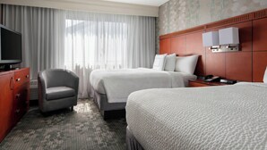 Quarto de hotel com duas camas, televisão, uma cadeira e mesa de cabeceira com telefone e controle remoto.