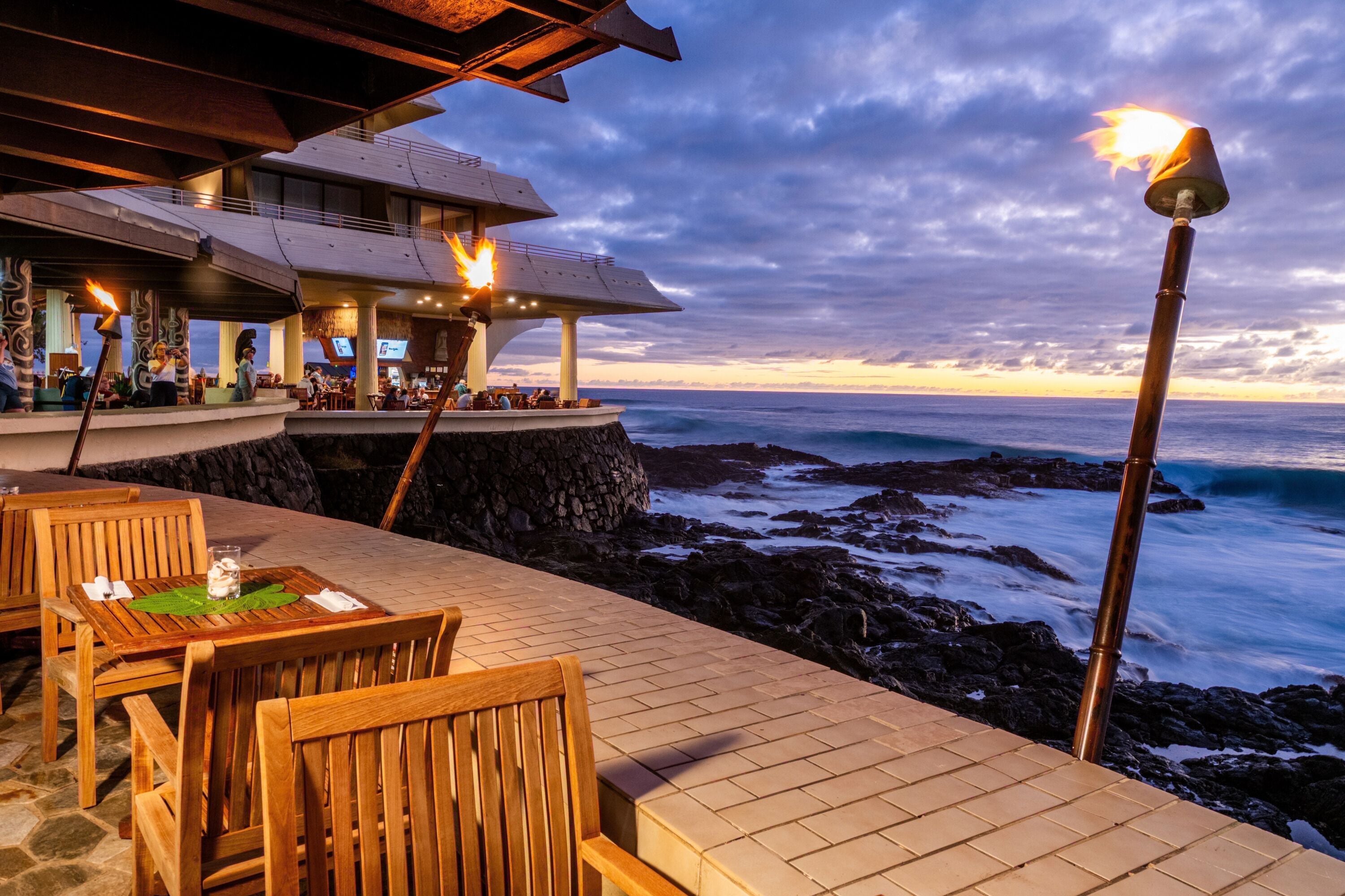 Restaurante à beira-mar com área externa, parede de pedra e vista para o oceano.