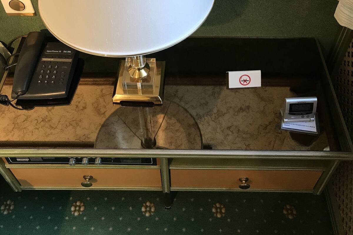 Quarto de hotel com escrivaninha, lâmpada, telefone, um pequeno dispositivo eletrônico e placa indicando proibido fumar.