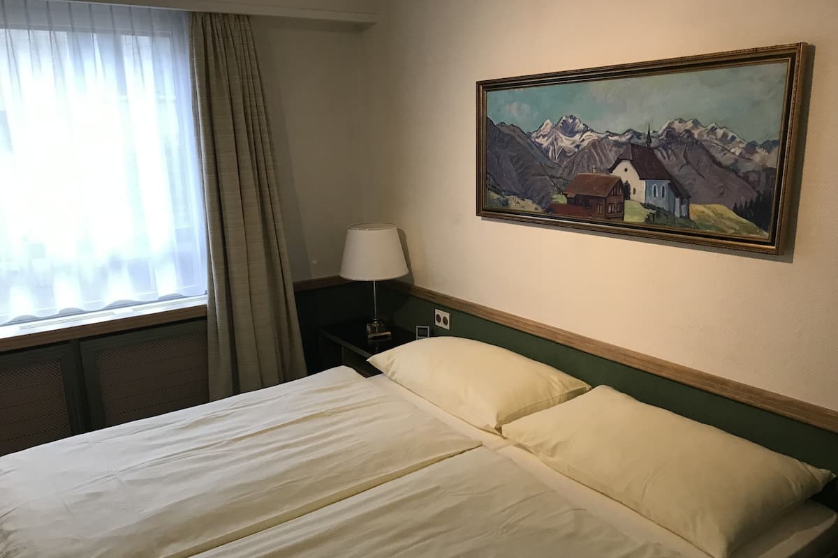 Quarto de hotel com cama, pintura de montanhas, janela com cortinas e mesa de cabeceira com abajur.