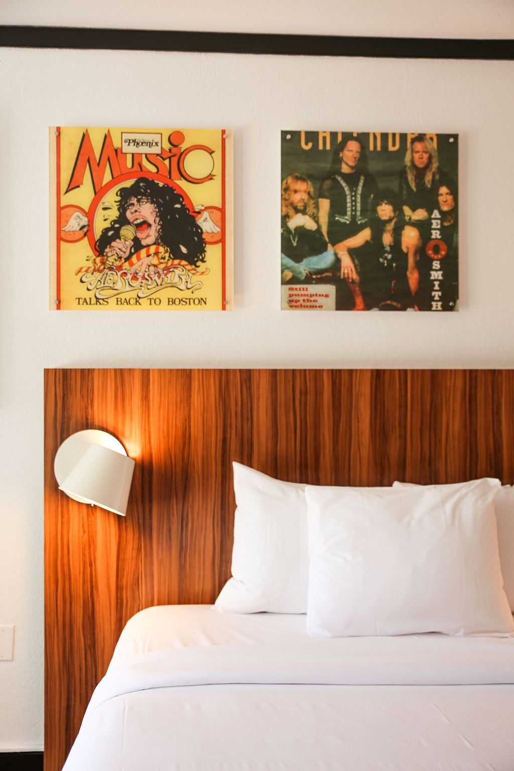 Quarto de hotel com uma cama, uma luminária fixada na parede e dois cartazes.