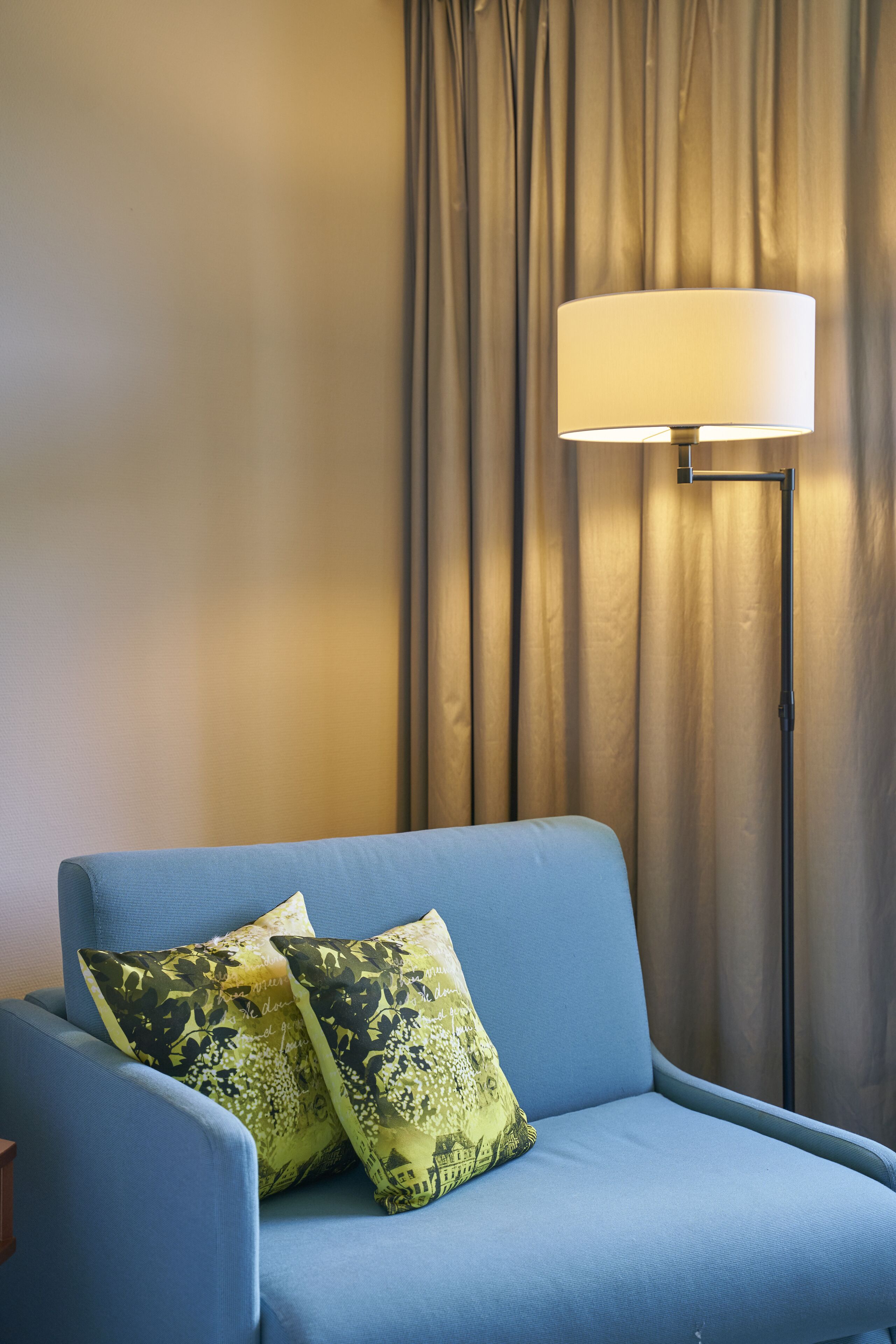 Um sofá azul com almofadas estampadas, uma luminária de chão e um fundo de cortina.