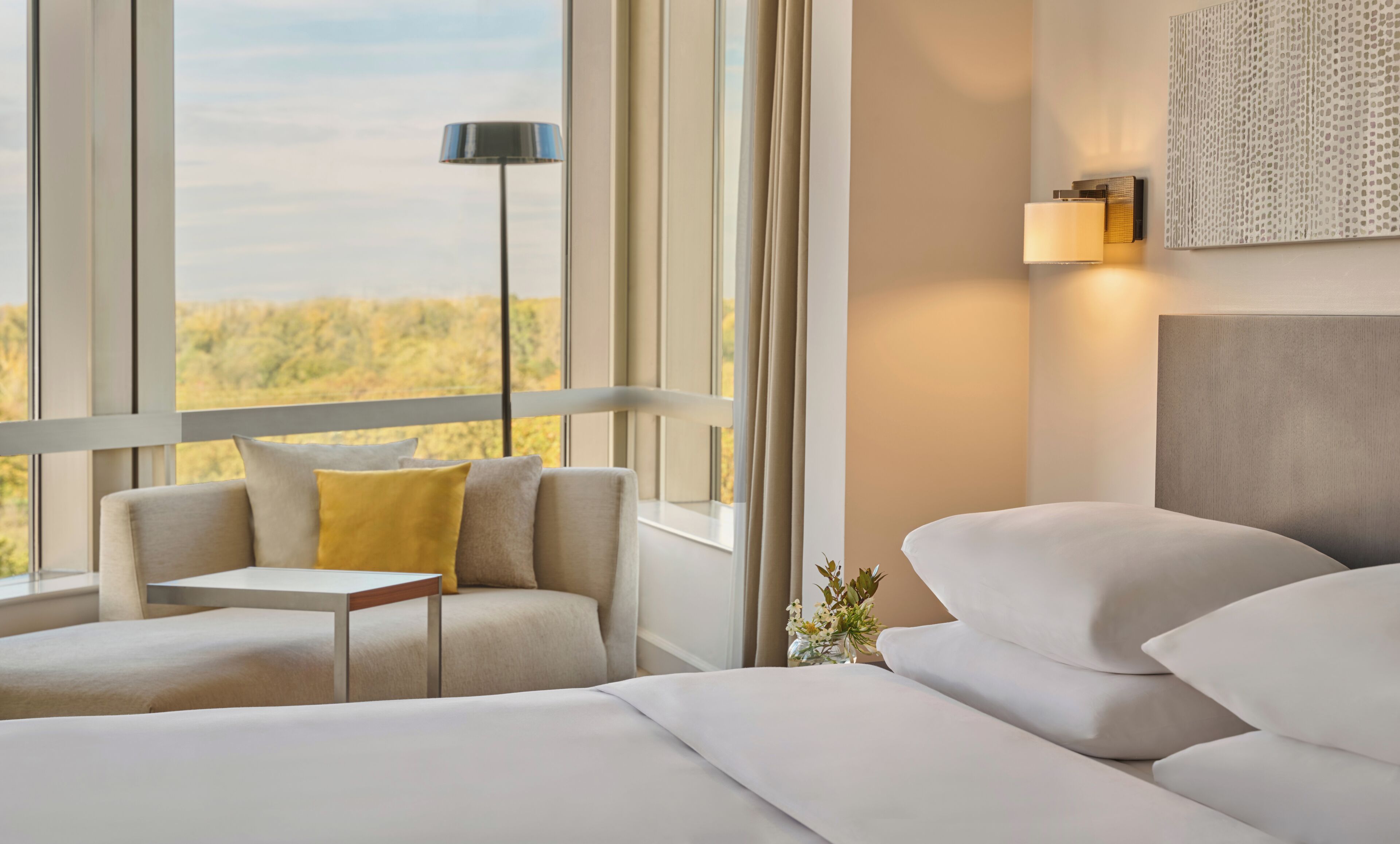 Quarto de hotel com cama, sofá, mesa pequena, abajur e vista para o exterior.