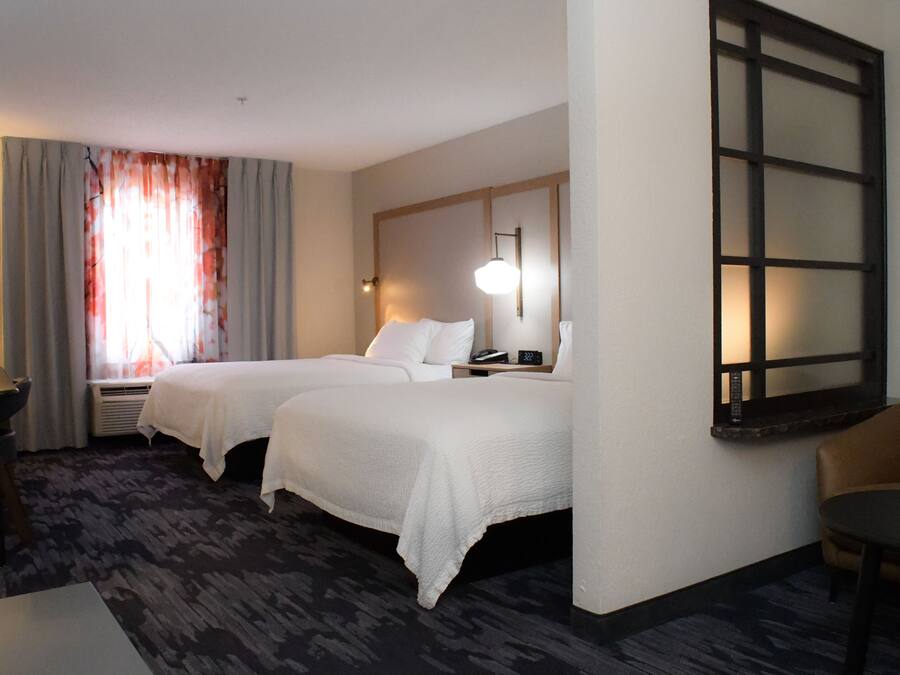 Quarto de hotel com duas camas, uma escrivaninha, uma cadeira e uma janela grande com cortinas.