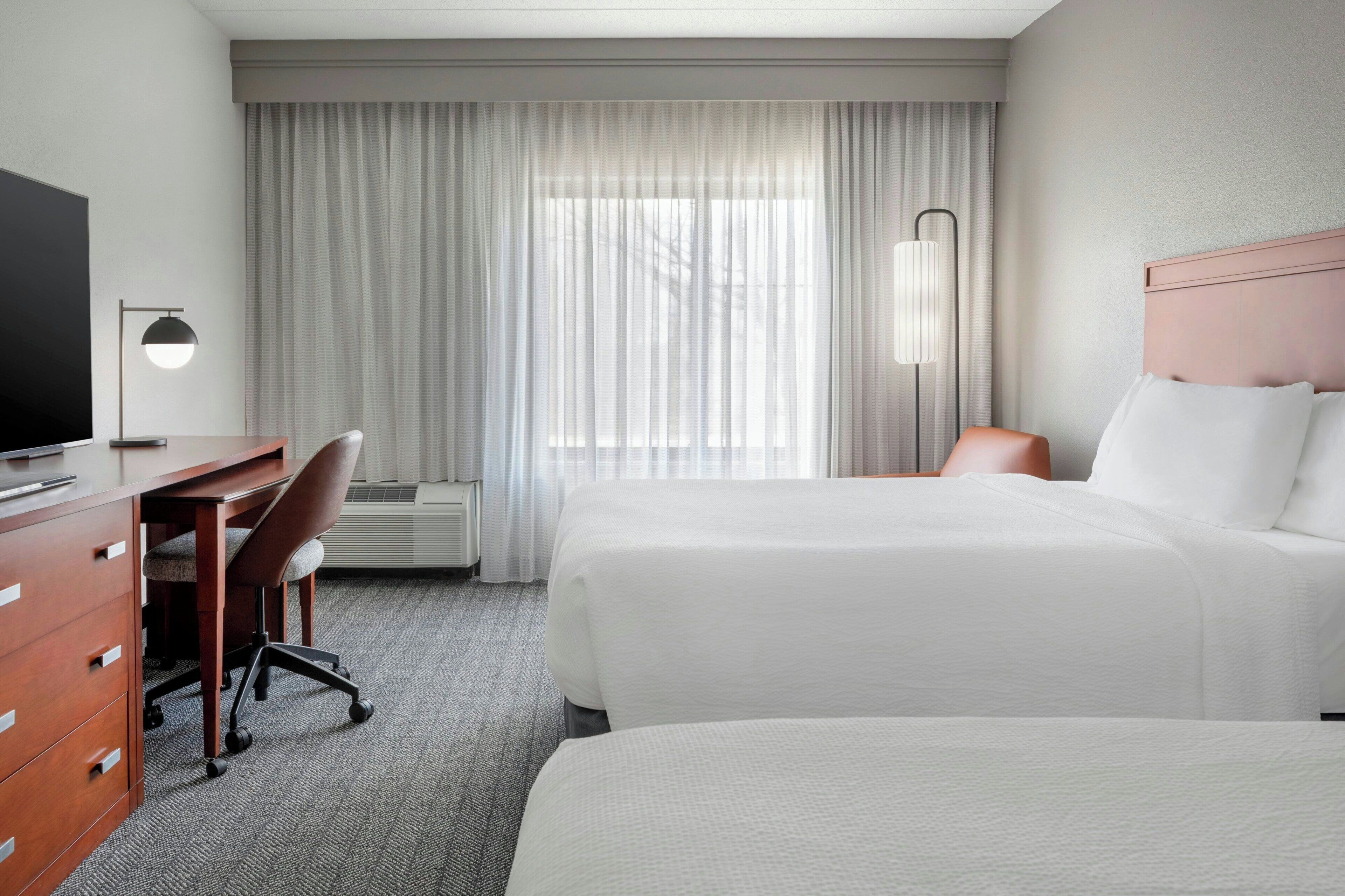 Quarto de hotel com cama, escrivaninha, cadeira, televisão e luminária.