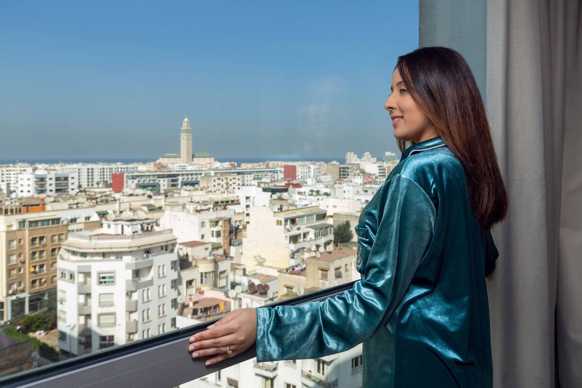 Uma pessoa em pé perto de uma janela com vista para um horizonte urbano com diversos edifícios e uma torre imponente.