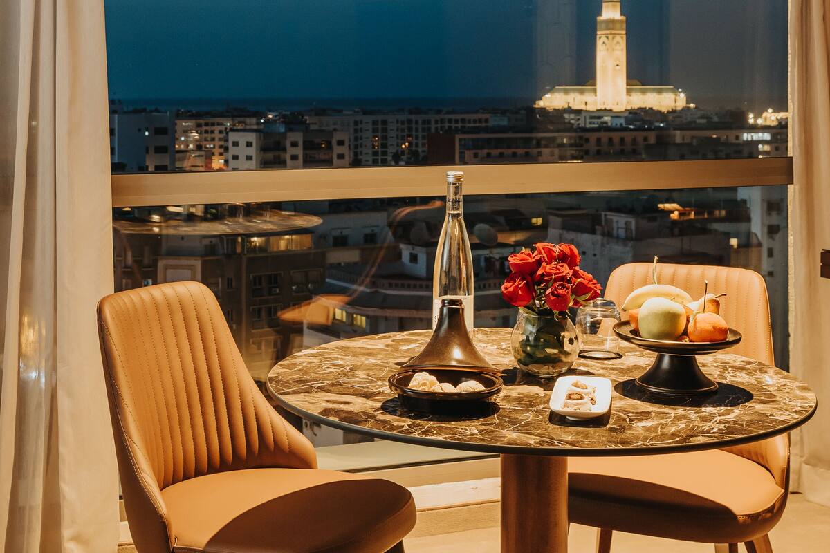 Uma sala de jantar com uma mesa posta para dois, um vaso de rosas vermelhas e vista para um horizonte urbano com uma torre imponente.
