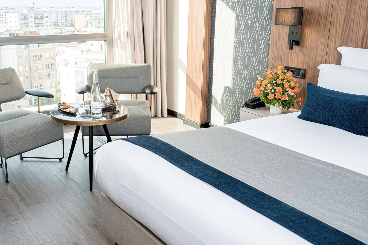Quarto de hotel com cama, duas poltronas, uma mesinha com garrafa e copos, um vaso com flores e vista para a cidade pela janela.