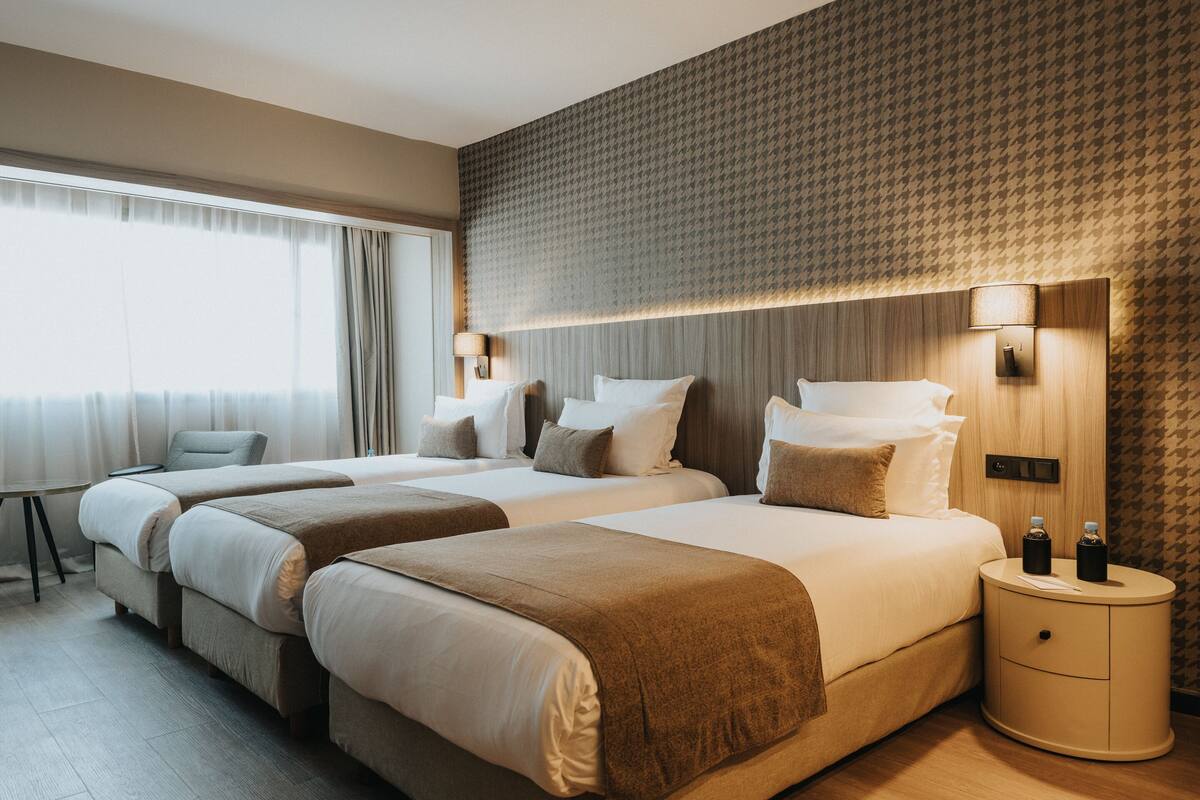 Quarto de hotel com três camas, uma escrivaninha e uma mesa de cabeceira com abajur.