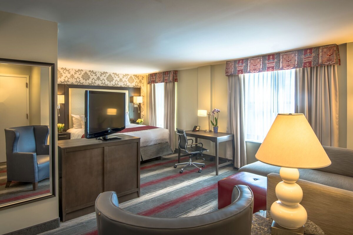 Quarto de hotel com cama, escrivaninha, cadeira, televisão, luminária e vista para a cidade.