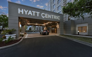 Entrada do hotel Hyatt Centric com design moderno, um carro estacionado na entrada e céu claro ao fundo.