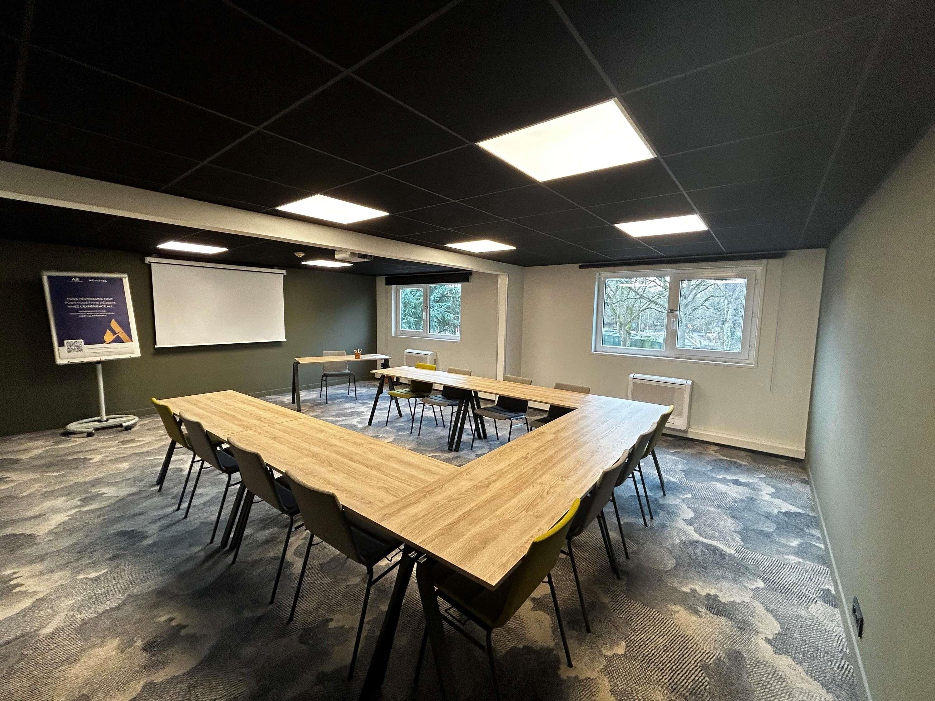 Uma sala de conferências com uma mesa comprida, cadeiras, uma tela de projetor e janelas.