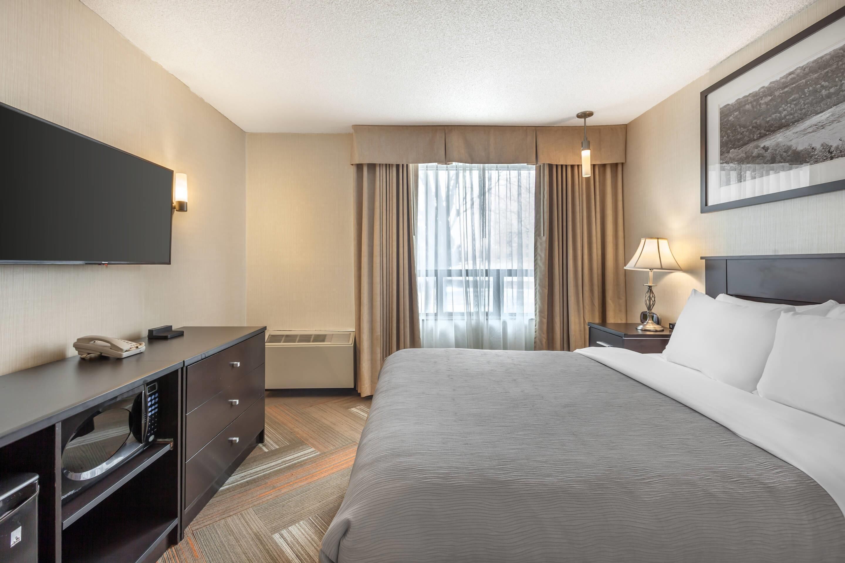 Quarto de hotel com cama, televisão, escrivaninha com telefone, luminária e janela com cortinas.